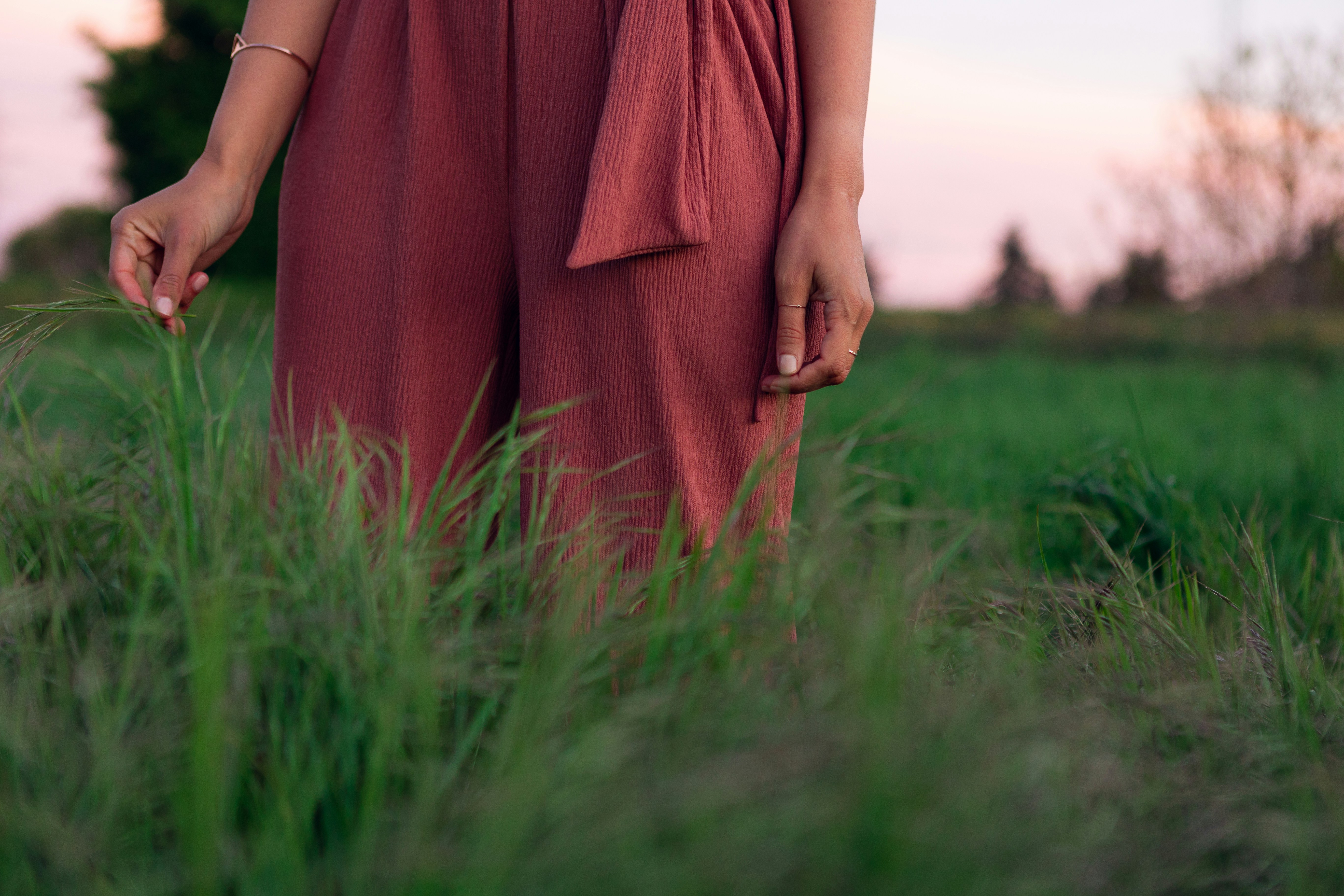 A person in stylish rust-colored pants gently touches the tall grass in a serene field during twilight. The soft colors of the sunset create a tranquil atmosphere.