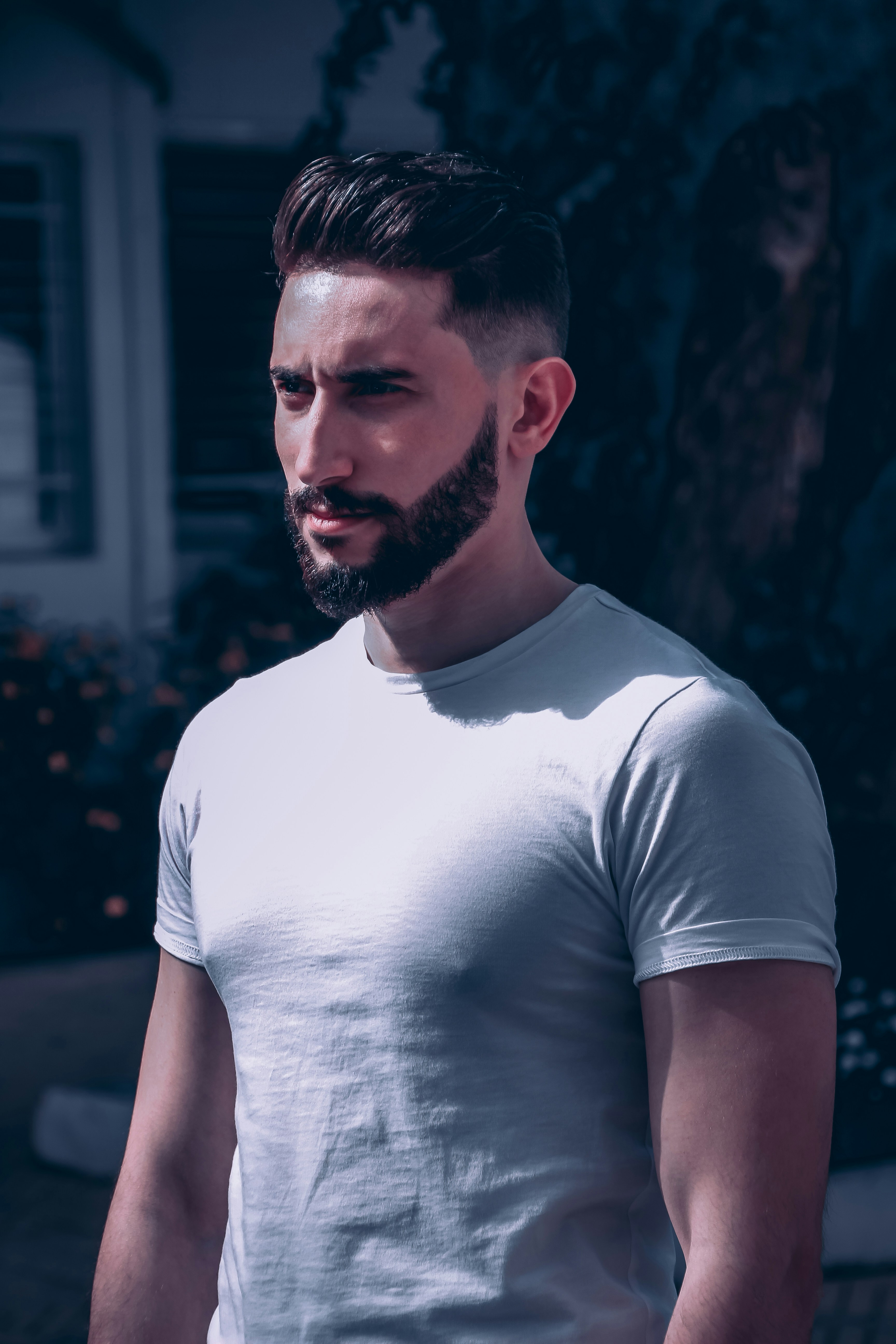 A man in a light gray t-shirt stands thoughtfully against a backdrop of greenery and soft sunlight, capturing a moment of introspection.