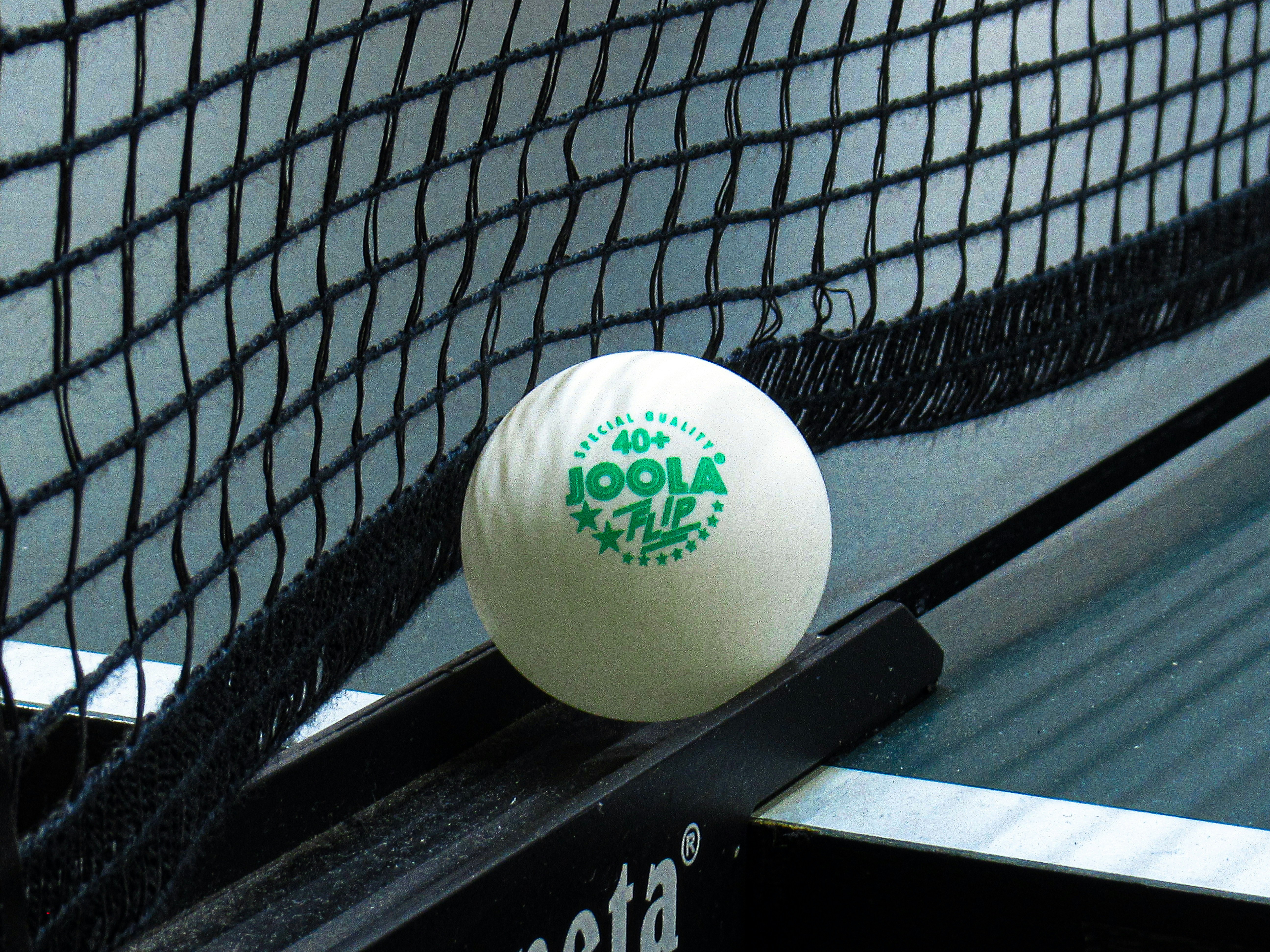 A white JOOLA ping-pong ball with a green JOOLA 40+ logo rests on the table edge near the net. The close-up highlights its smooth surface and the texture of the table.