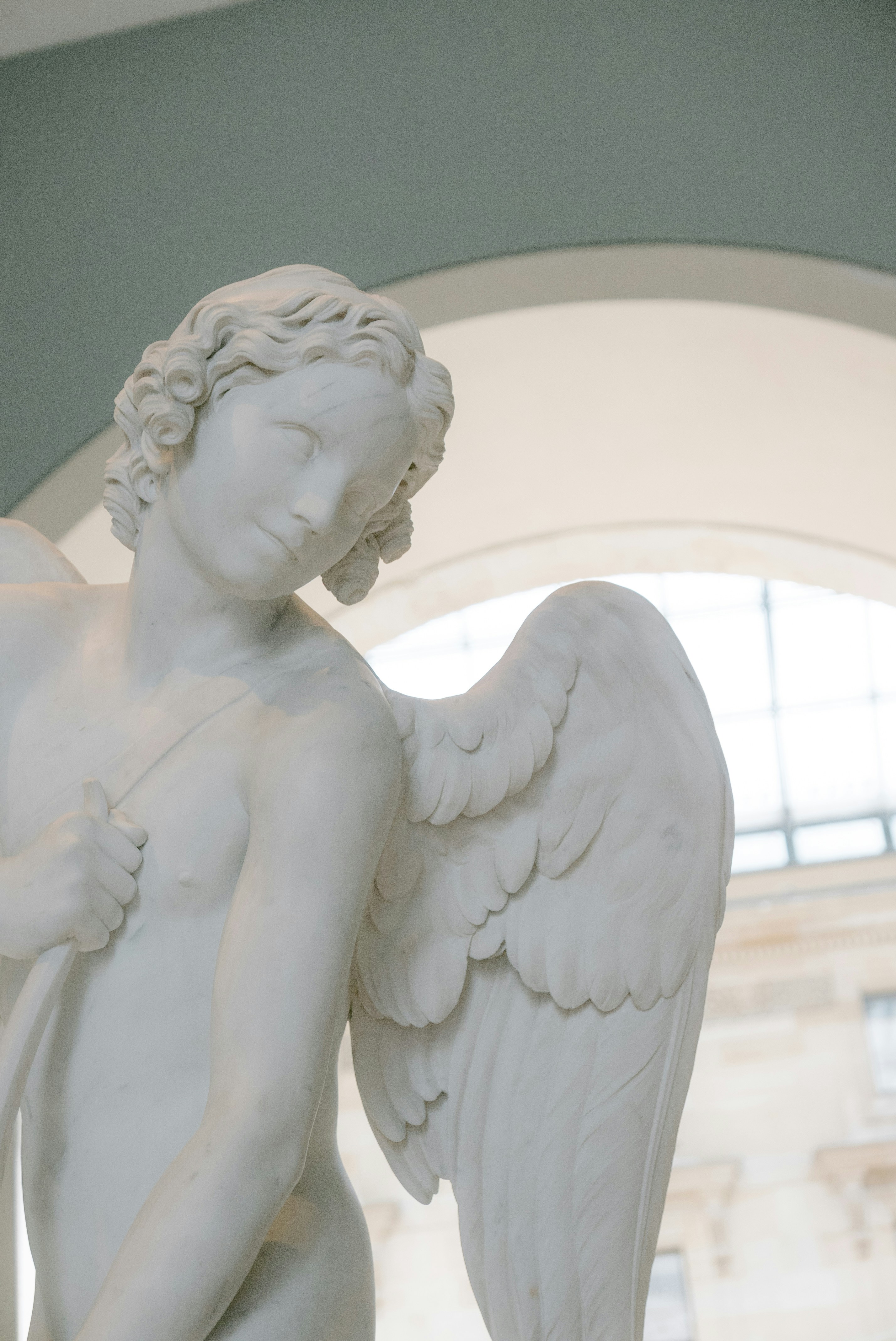 Elegant white marble sculpture of an angel, showcasing intricate details in the wings and facial expression, set against a softly lit architectural backdrop.