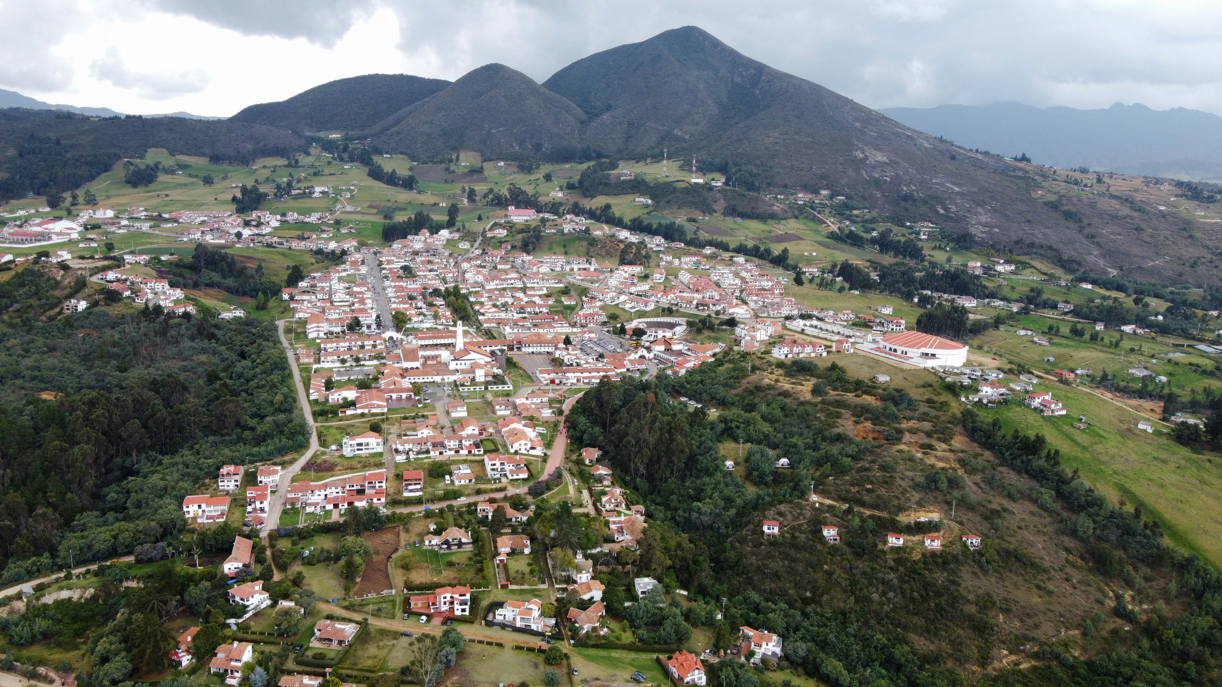 Aerial view of Guatavita's red-roofed town set against lush greenery and rolling hills under a cloudy sky.