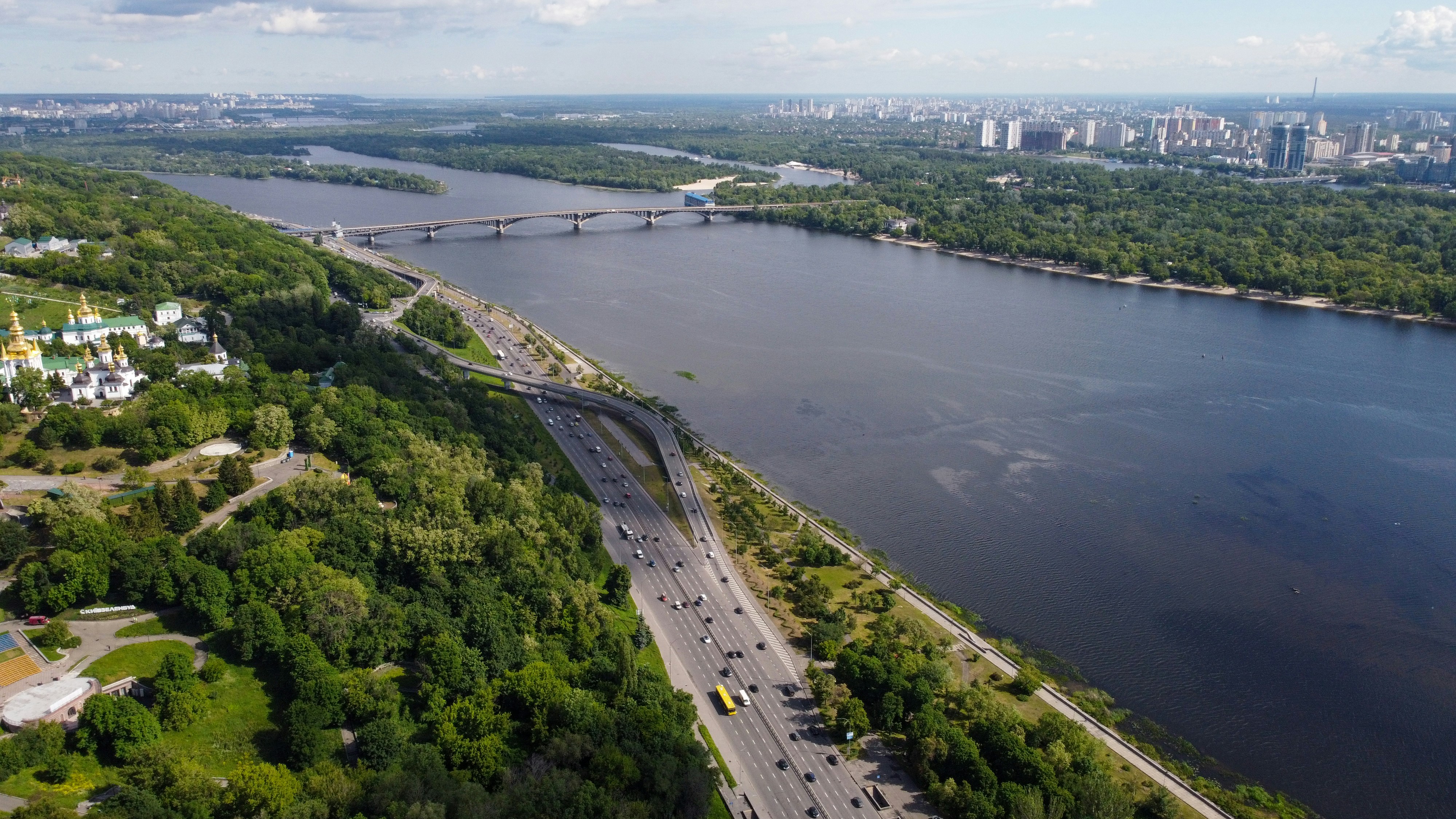 Aerial view showcasing a river winding through a lush landscape, with a busy highway and city skyline in the distance.