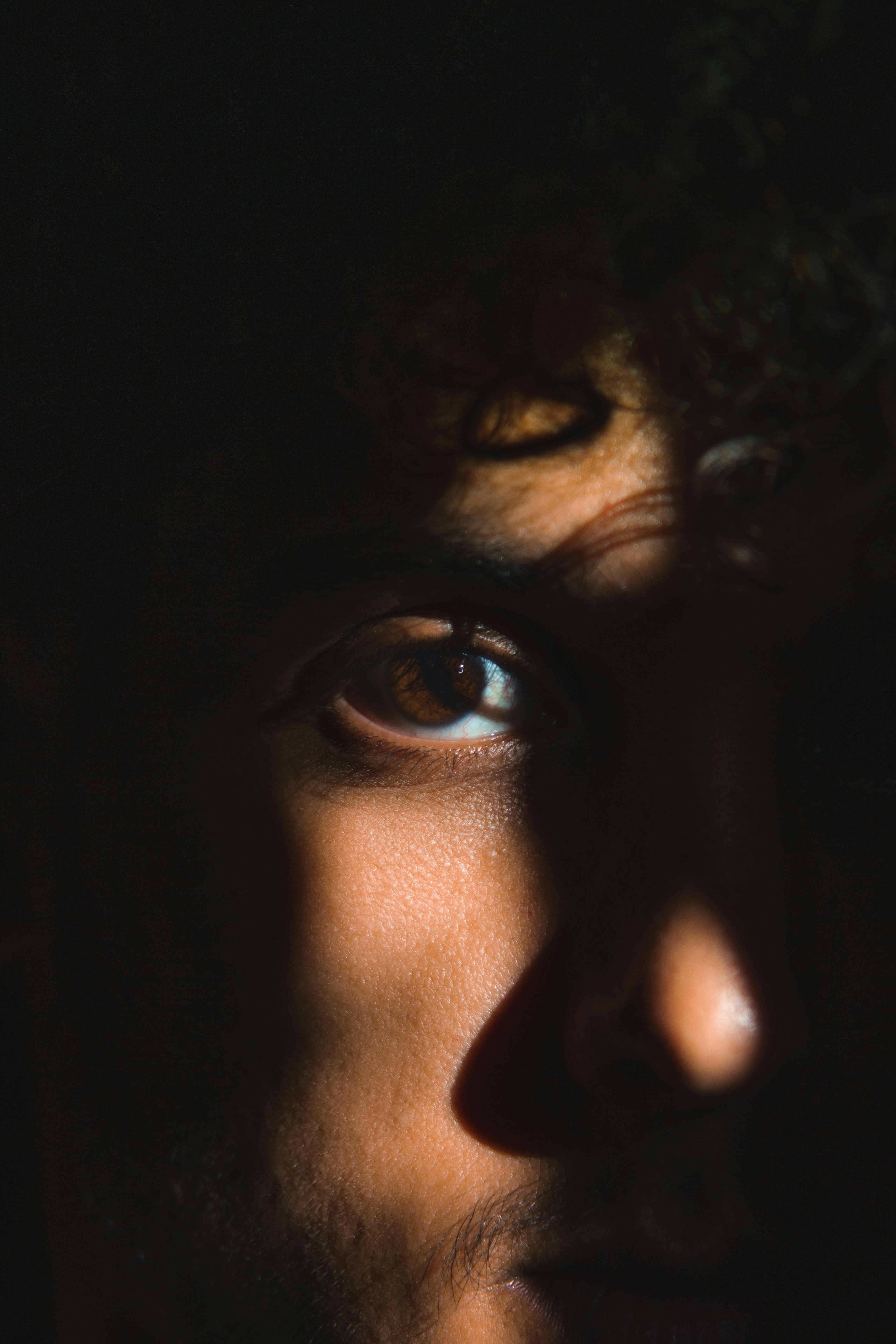Close-up of a person's eye partially illuminated by light, with dramatic shadows creating a striking contrast. The image captures an intense moment of introspection.