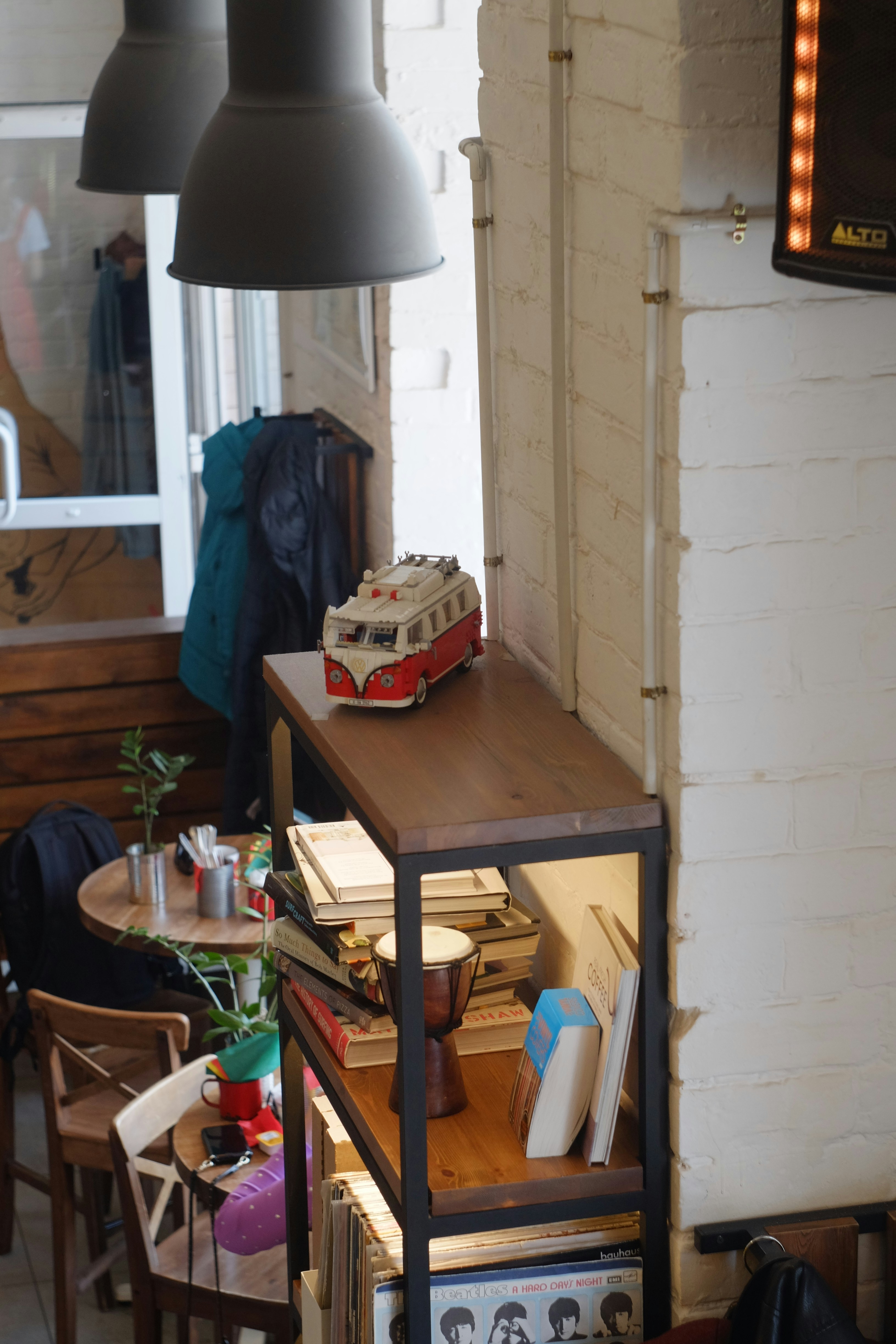 A vintage toy bus sits atop a wooden shelf filled with books and decorative items in a warm café setting.