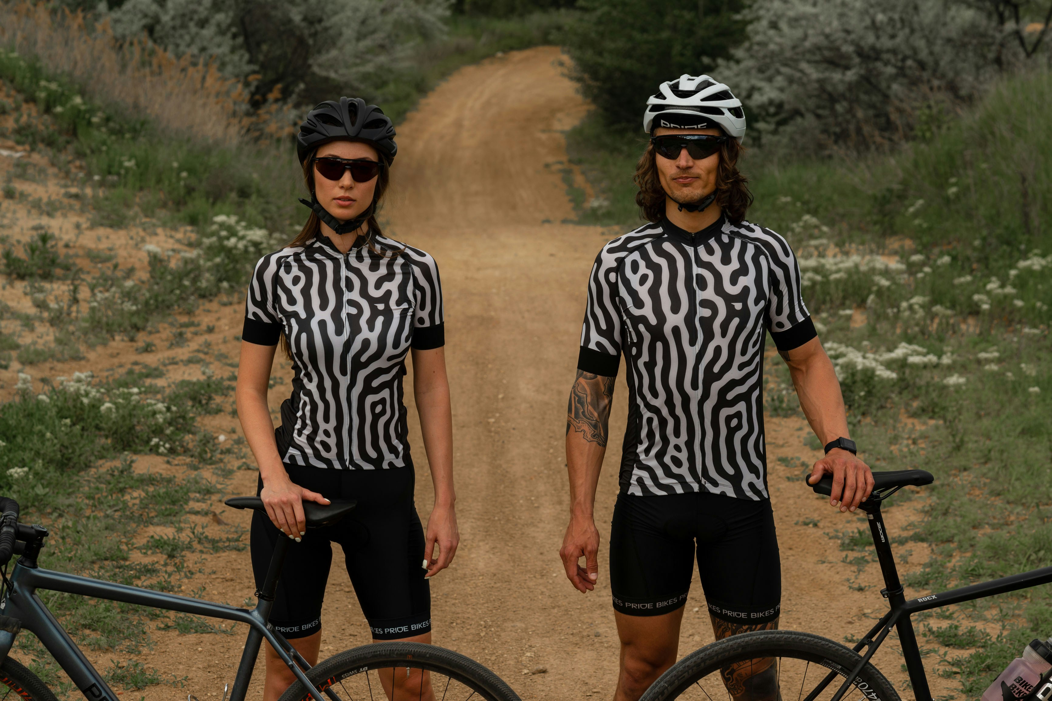 Two cyclists in zebra-pattern jerseys stand with their bikes on a dusty rural trail, facing the camera with helmets and sunglasses, ready for a ride.