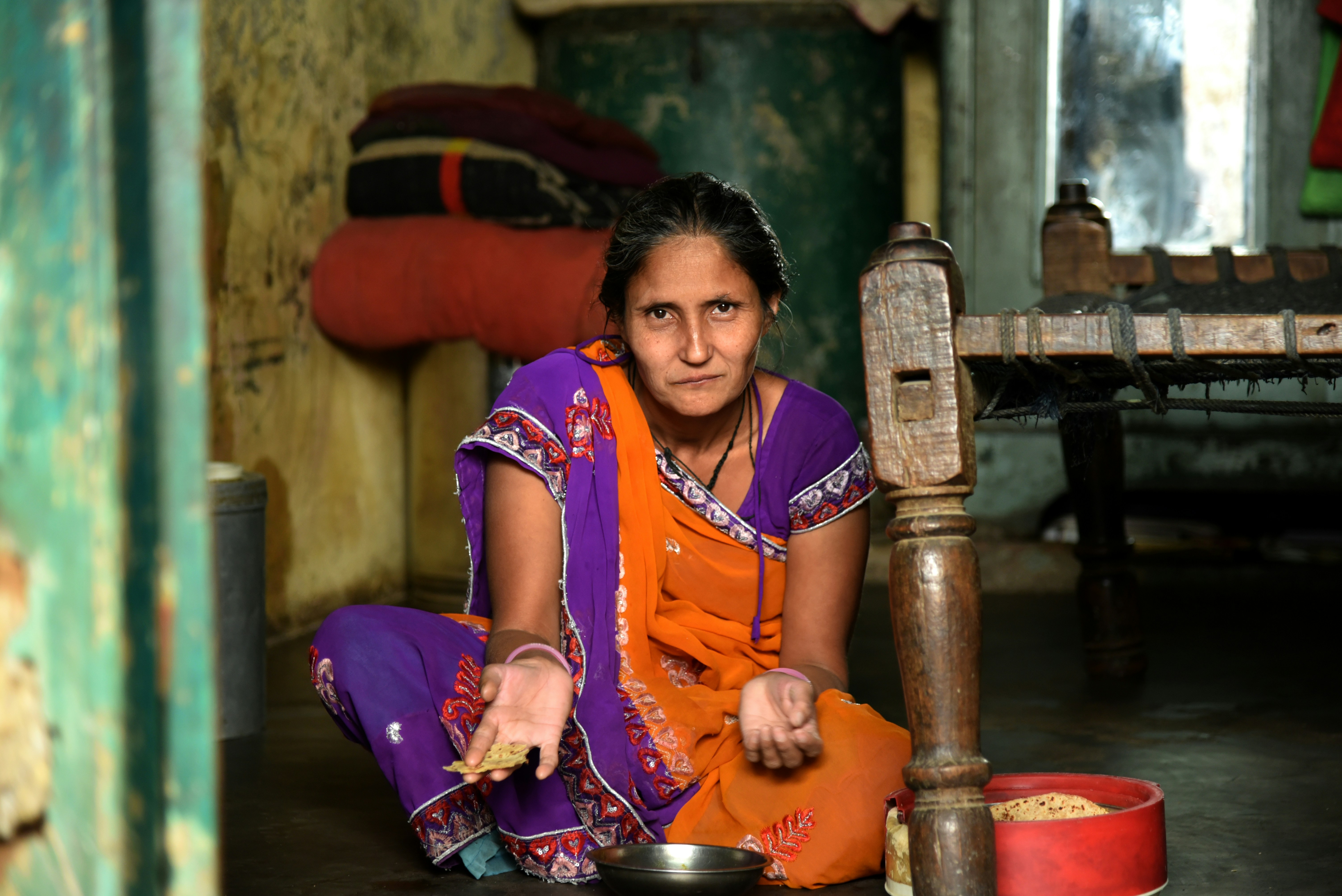 Woman in a colorful saree sitting on the floor with food in a rustic room.