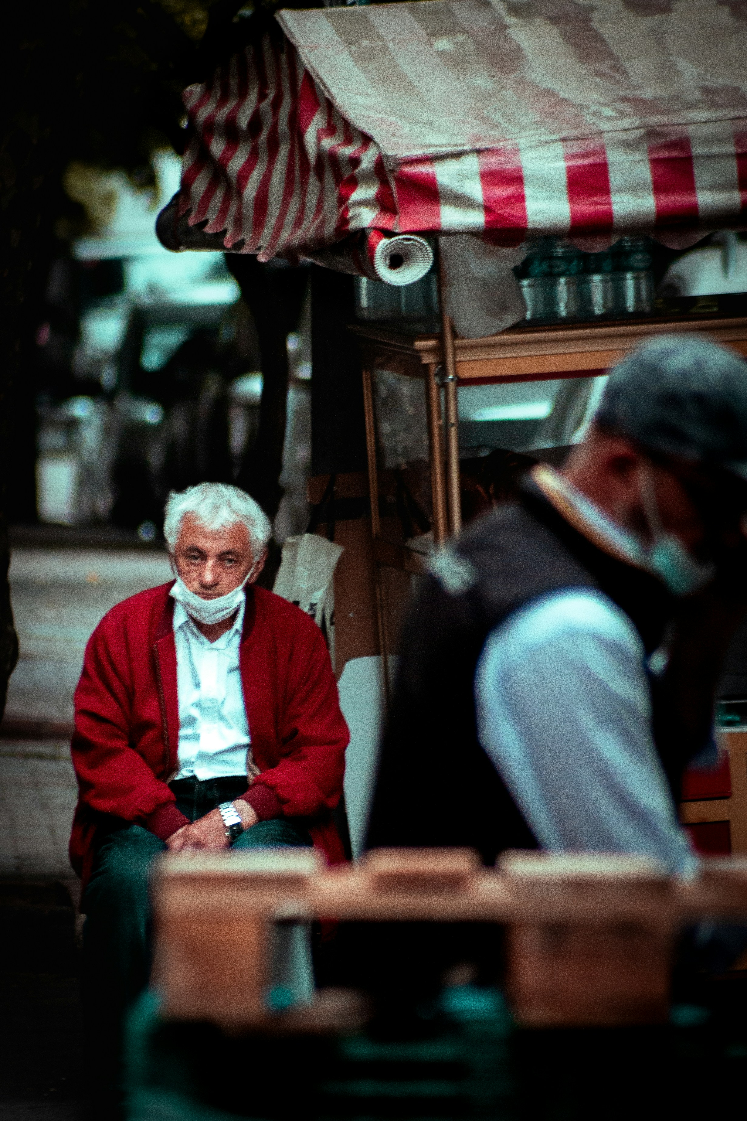 Paused Under the Striped Canopy: A Masked Elder by the Market Stall