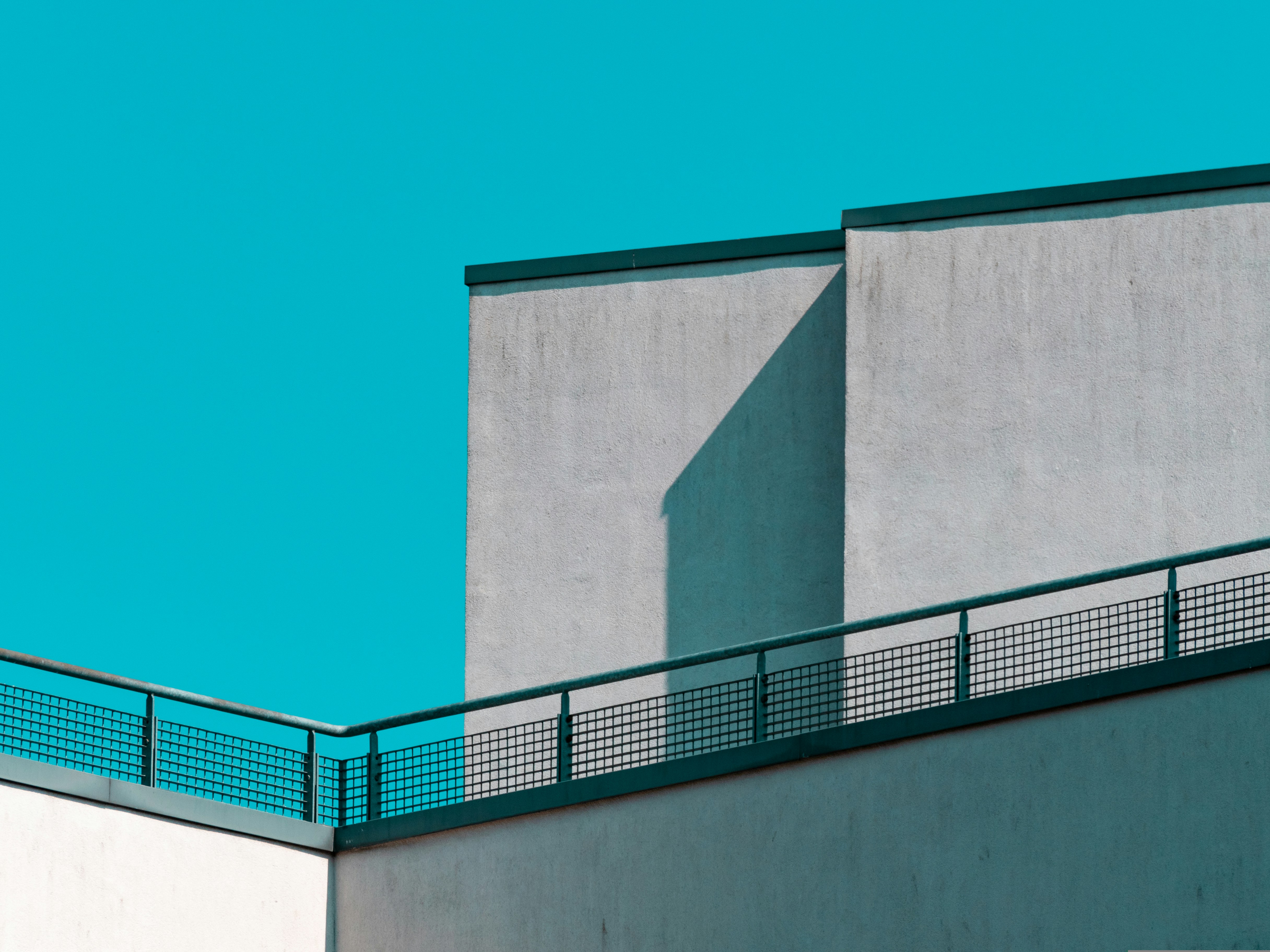 Minimalist rooftop scene with concrete walls, a metal railing, and a bold diagonal shadow set against a cyan sky.