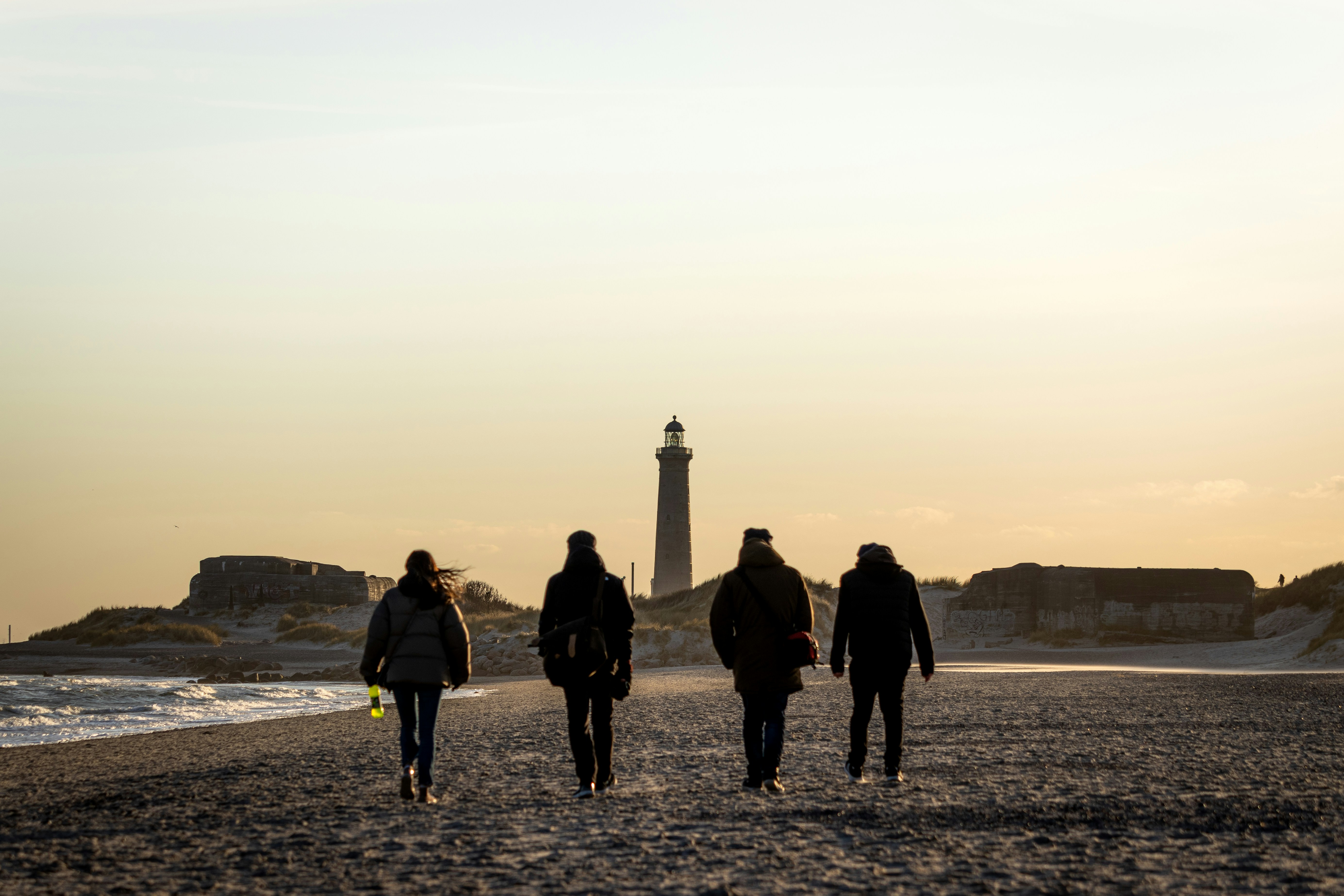 Four figures walking along a sandy beach towards a distant lighthouse under a soft sunset glow.