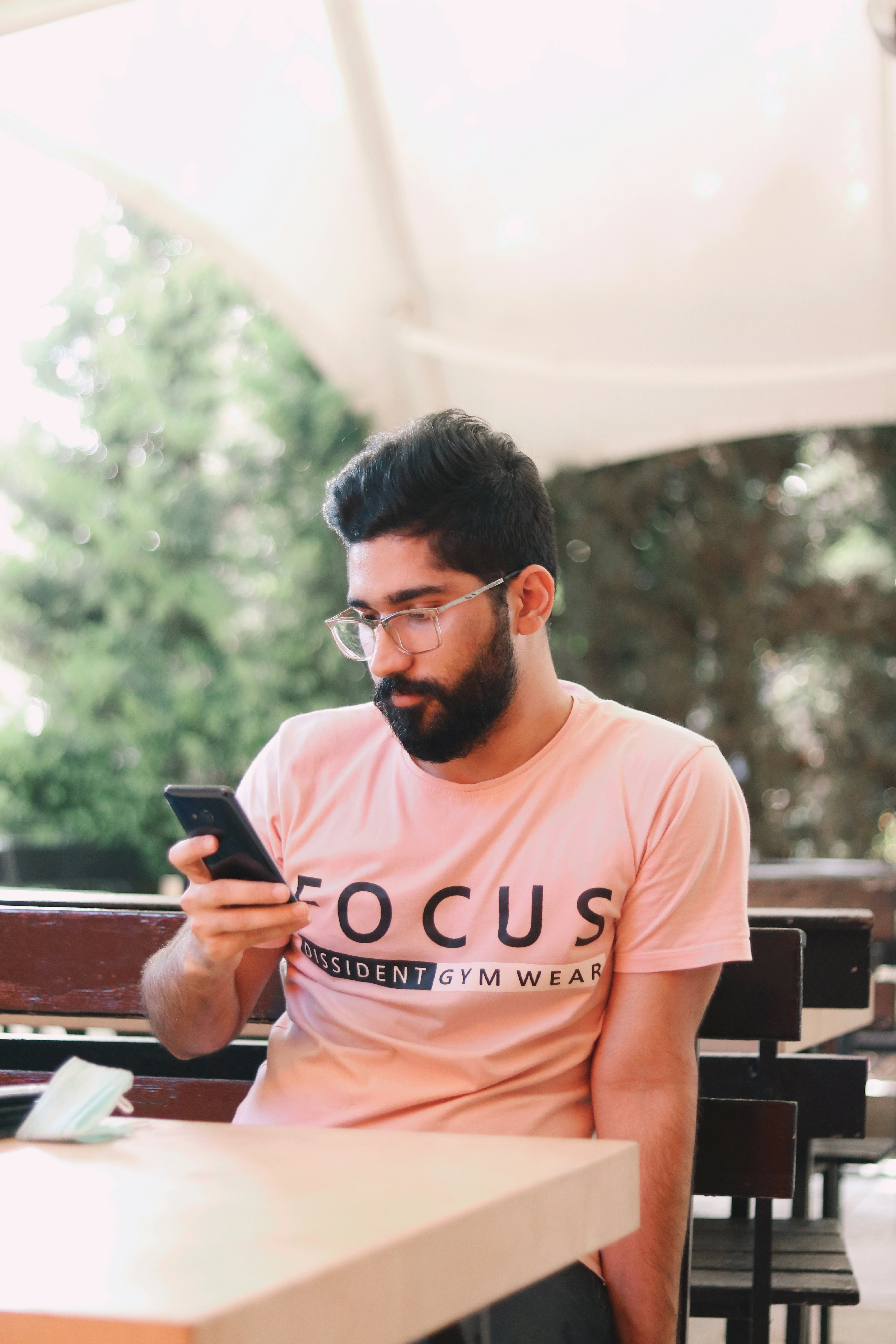 Young man in a pink shirt focused on his smartphone while seated at a café table, surrounded by greenery.