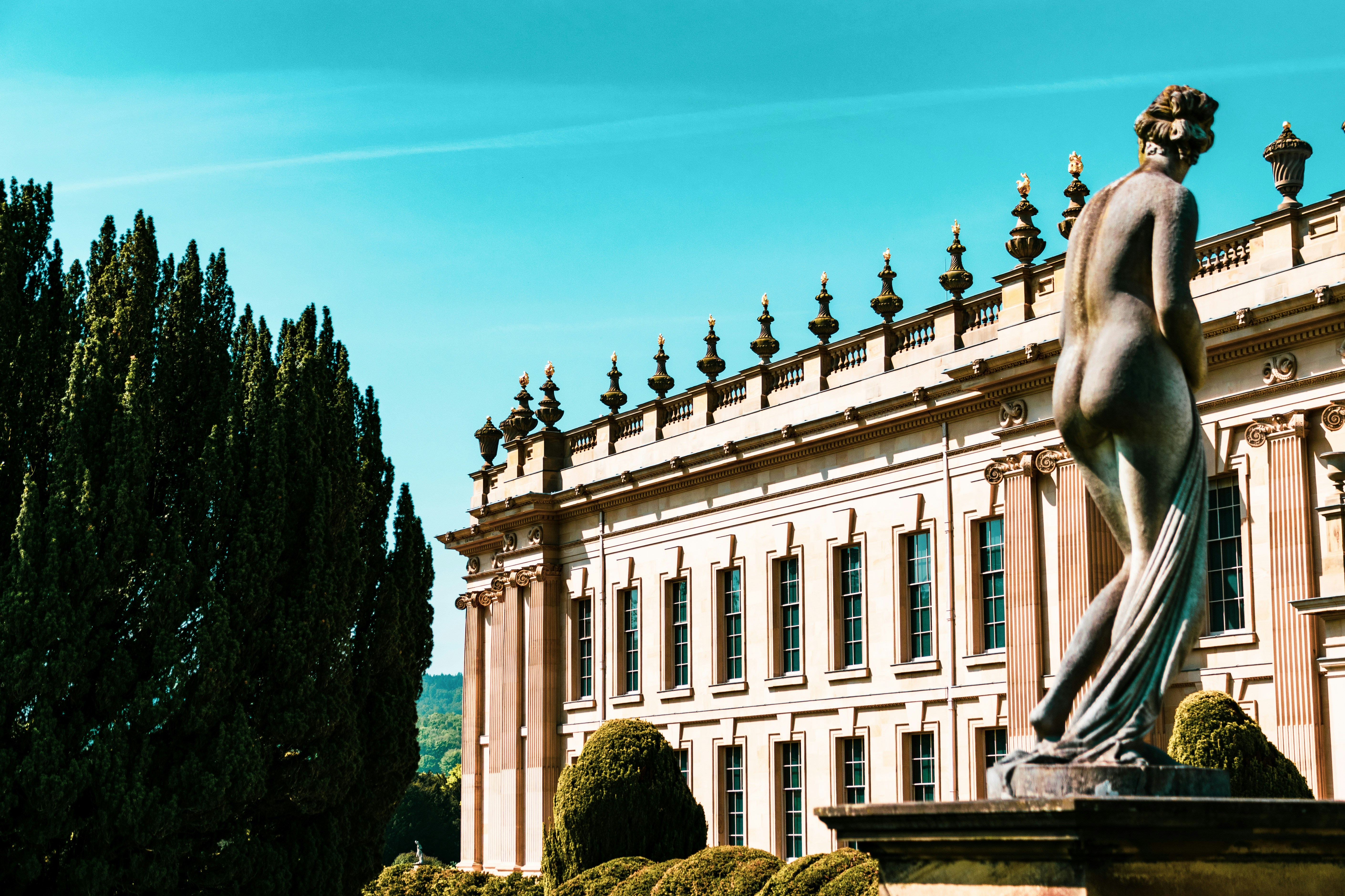 A classical statue stands gracefully in front of an ornate building, framed by lush greenery and a clear blue sky.