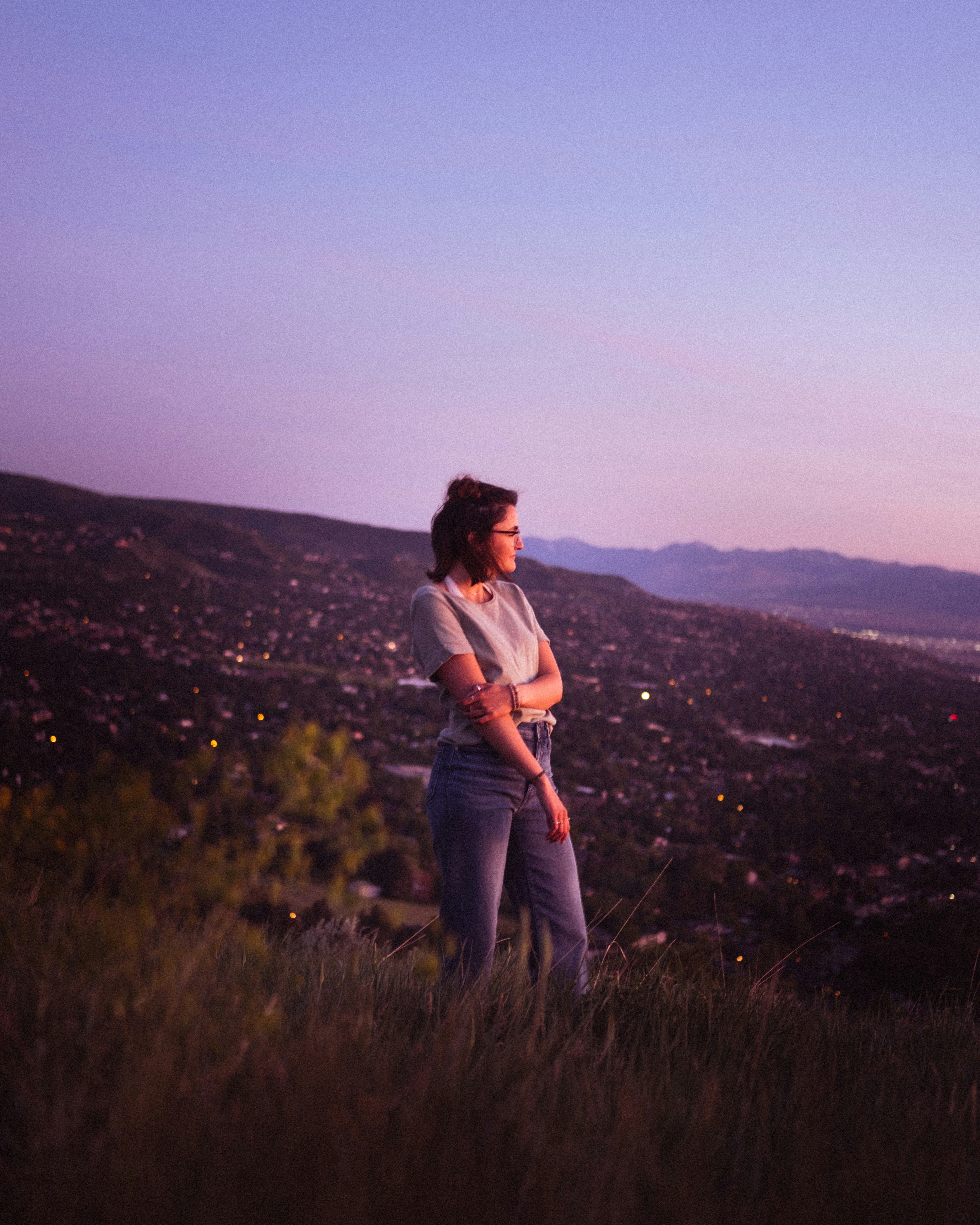Young woman standing on a hillside, gazing over a sprawling valley illuminated by city lights during twilight.