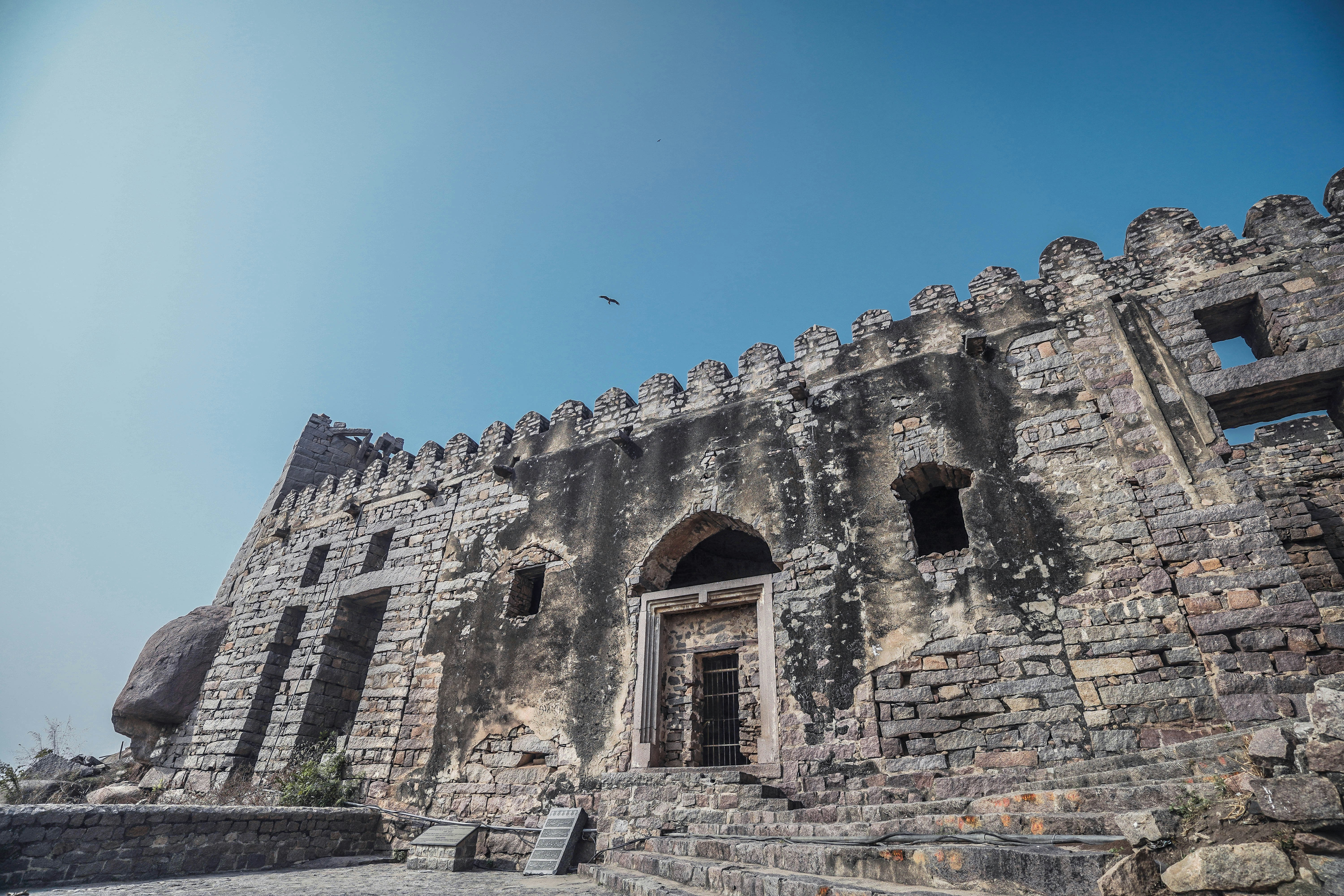 Ancient stone fortress with weathered walls and a prominent entrance, set against a clear blue sky. The structure reflects the passage of time and history.