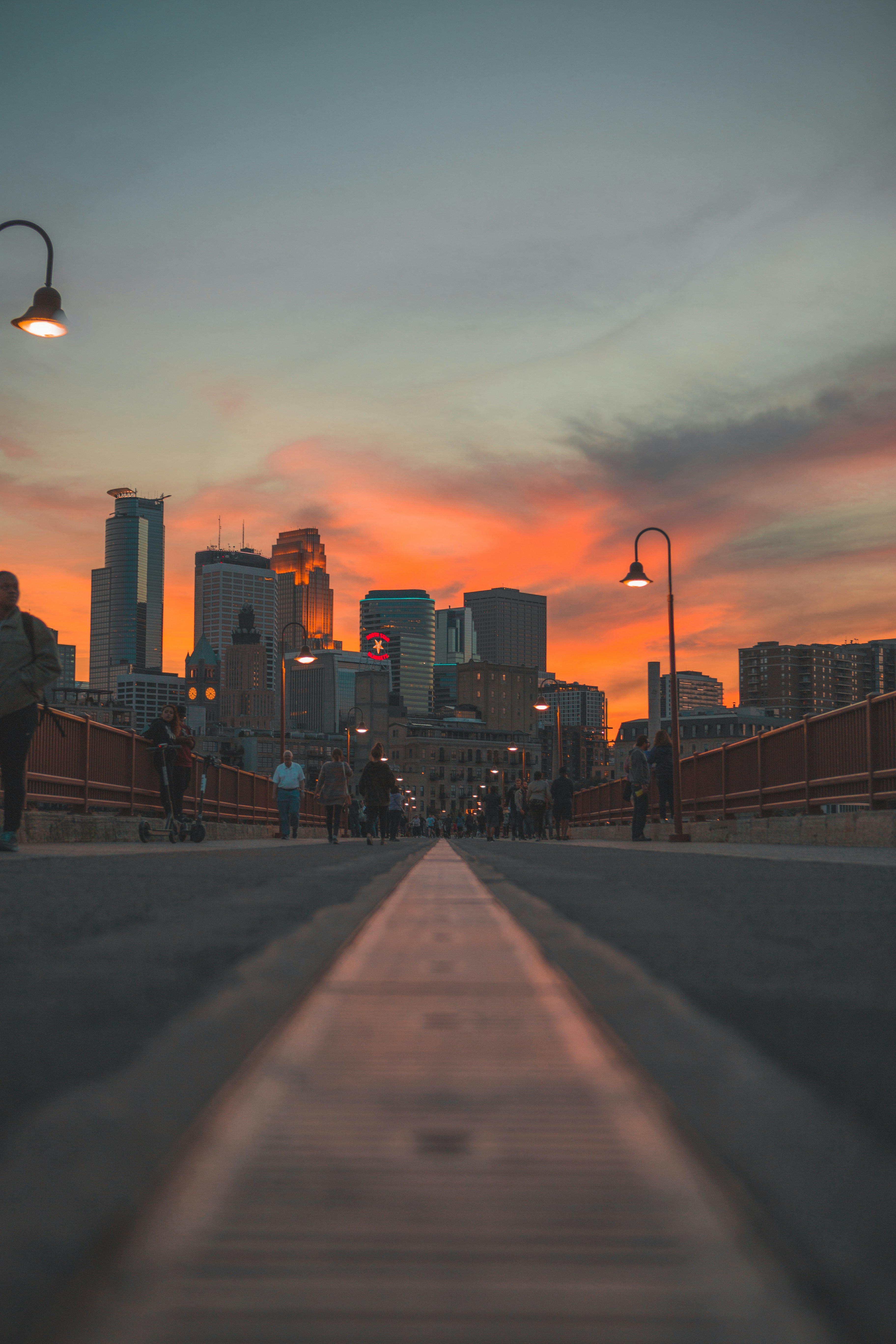 Pedestrians stroll along a bridge with a view of a vibrant sunset illuminating the city skyline. Street lamps cast a warm glow over the scene.