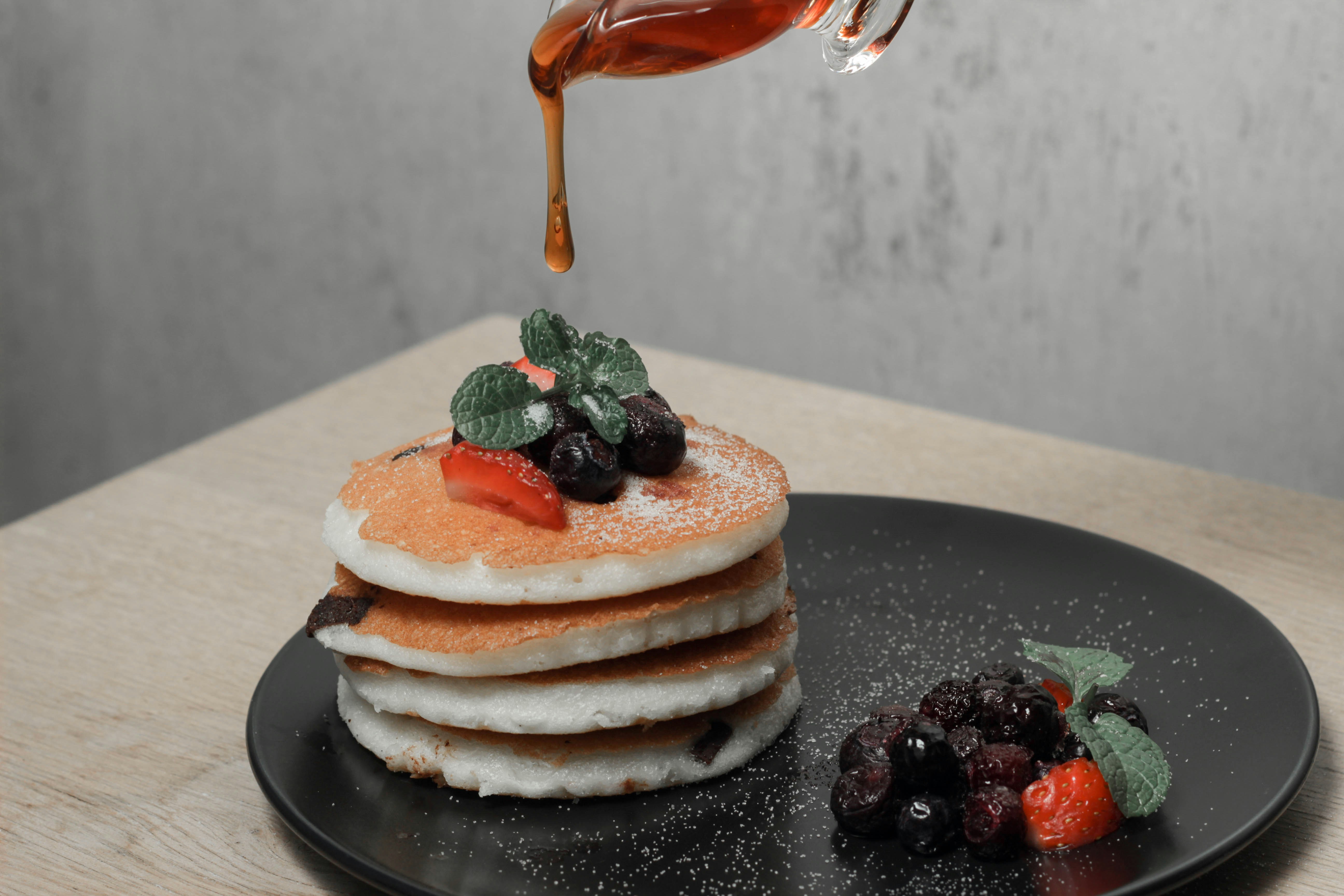 Fluffy pancakes stacked high, adorned with fresh berries and mint, as warm maple syrup cascades down. A delicious morning feast awaits.