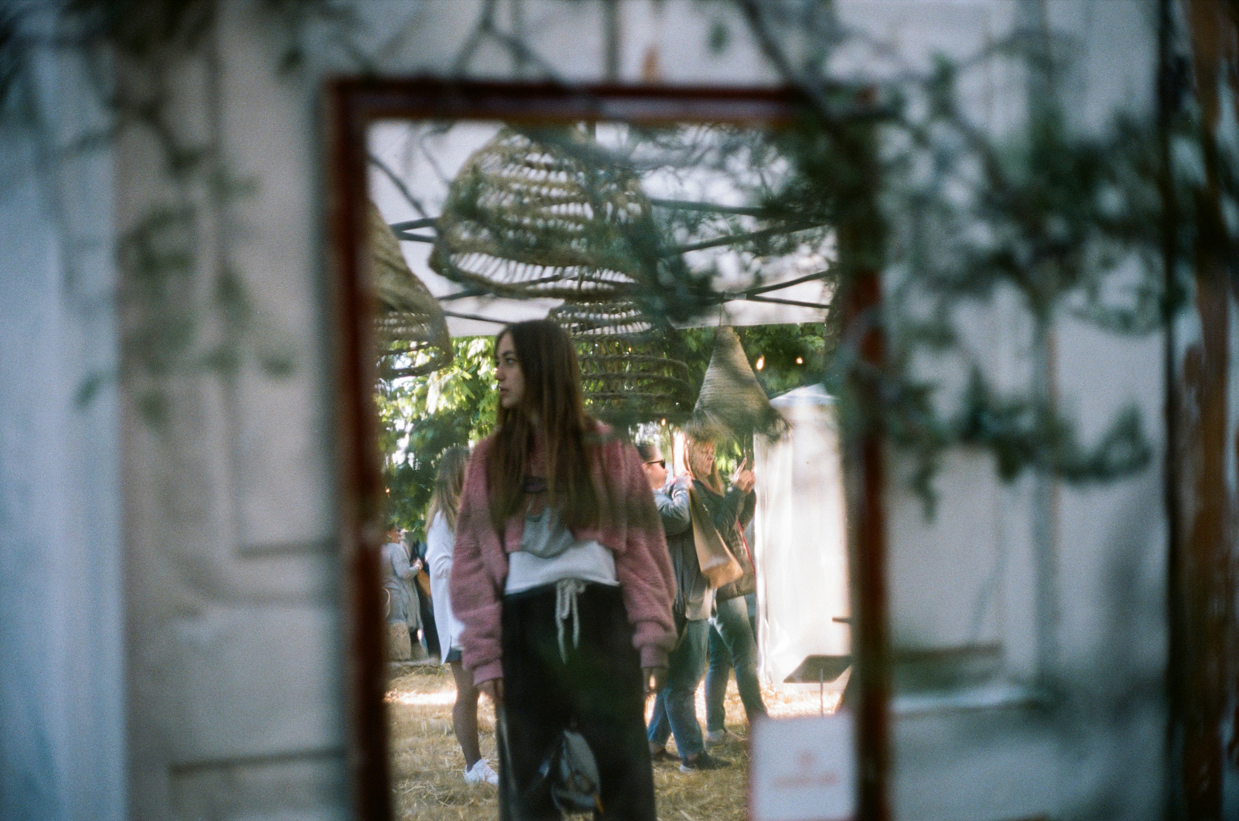 Photograph featuring a woman in a pink jacket standing in a doorway framed by greenery at an outdoor event, with blurred figures in the background.
