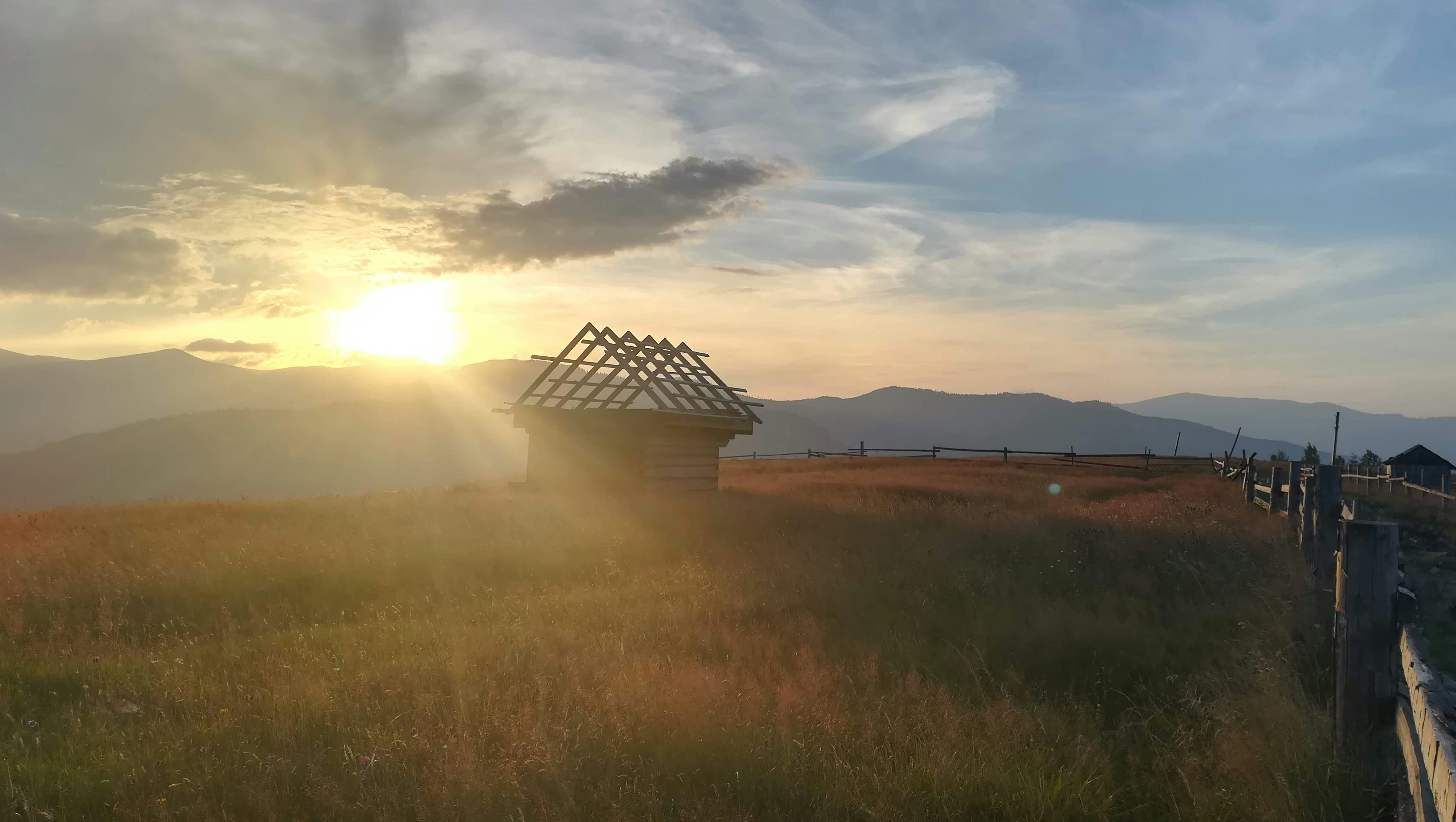 Golden sunlight spills over a rustic structure on a hillside, framed by distant mountains and a gentle fence. The scene captures the tranquility of sunset in a pastoral landscape.