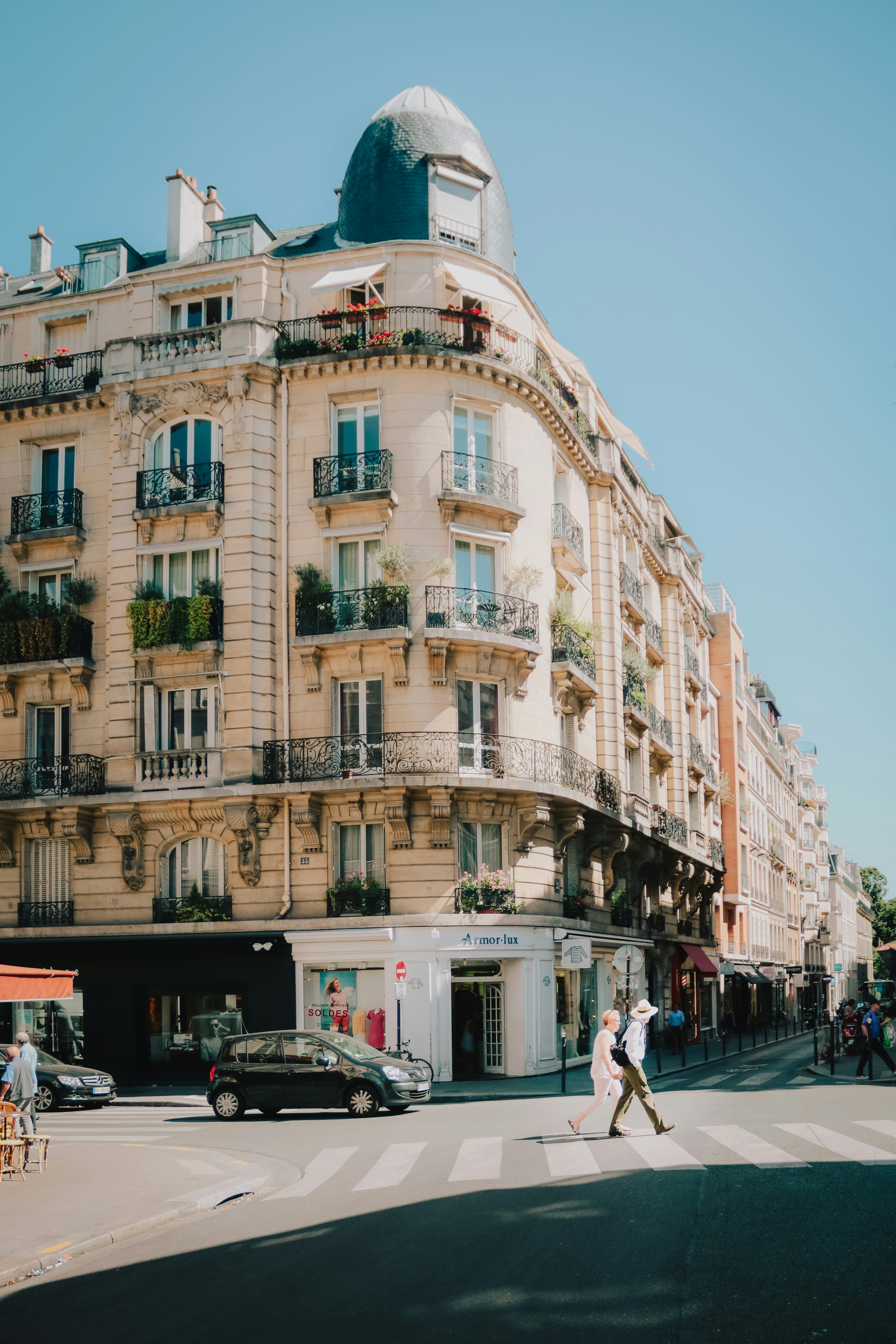 Charming Parisian architecture featuring a curved building with balconies, set against a clear blue sky. Pedestrians cross the street, adding life to the scene.