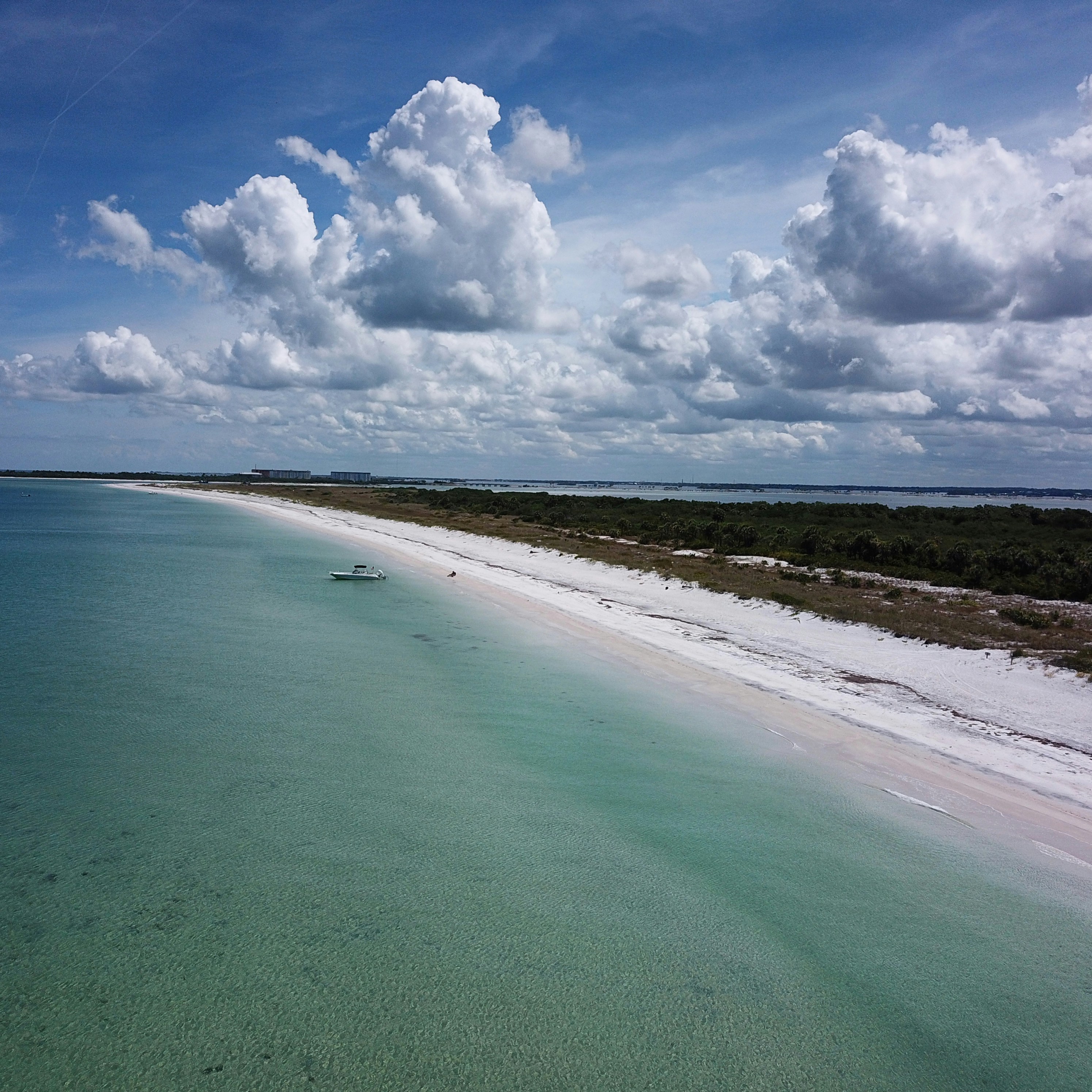 Gentle waves lap against a pristine beach, with a small boat anchored near the shore under a sky filled with fluffy clouds.