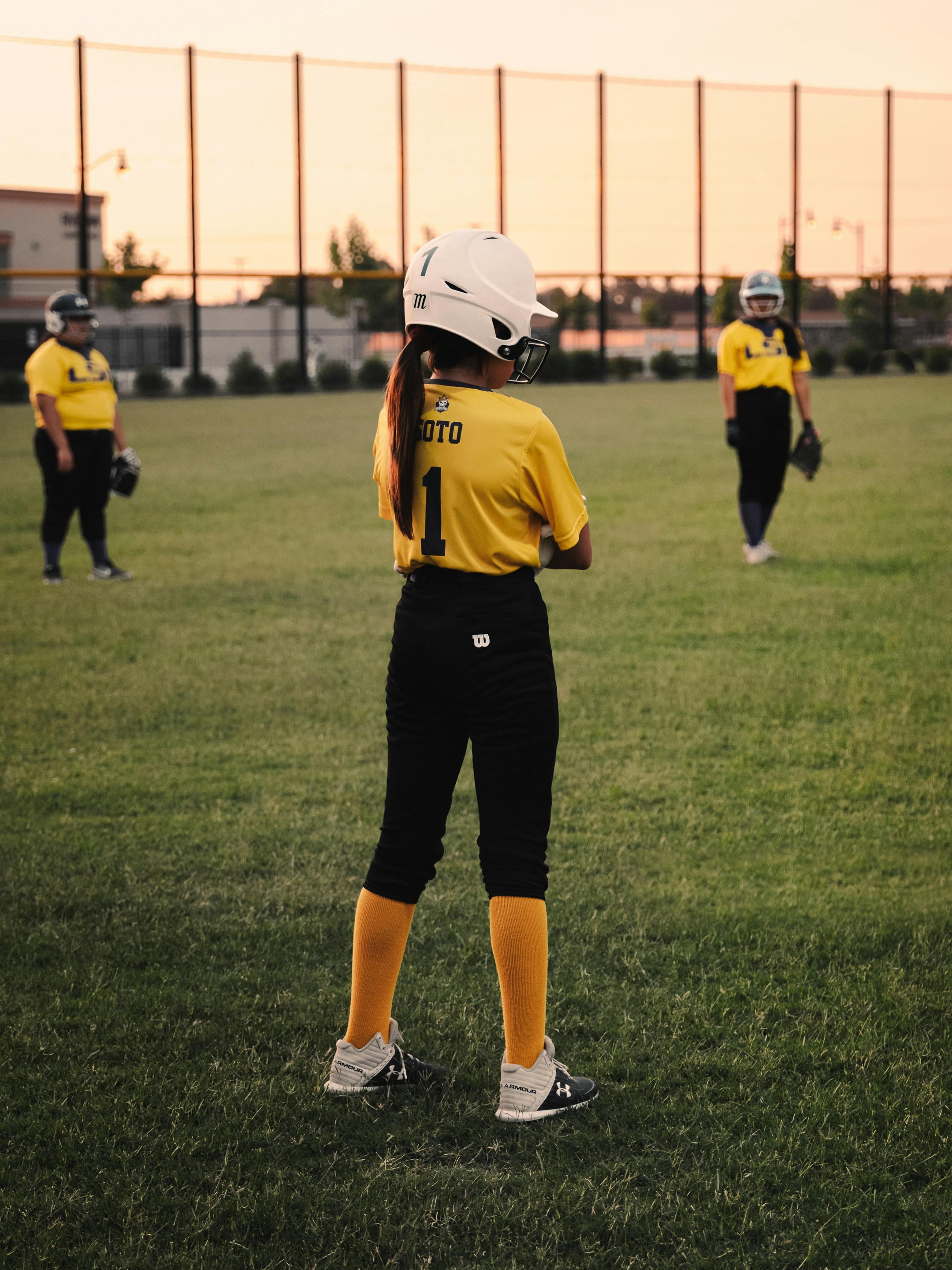 Softball players in yellow uniforms stand on a grassy field at sunset.
