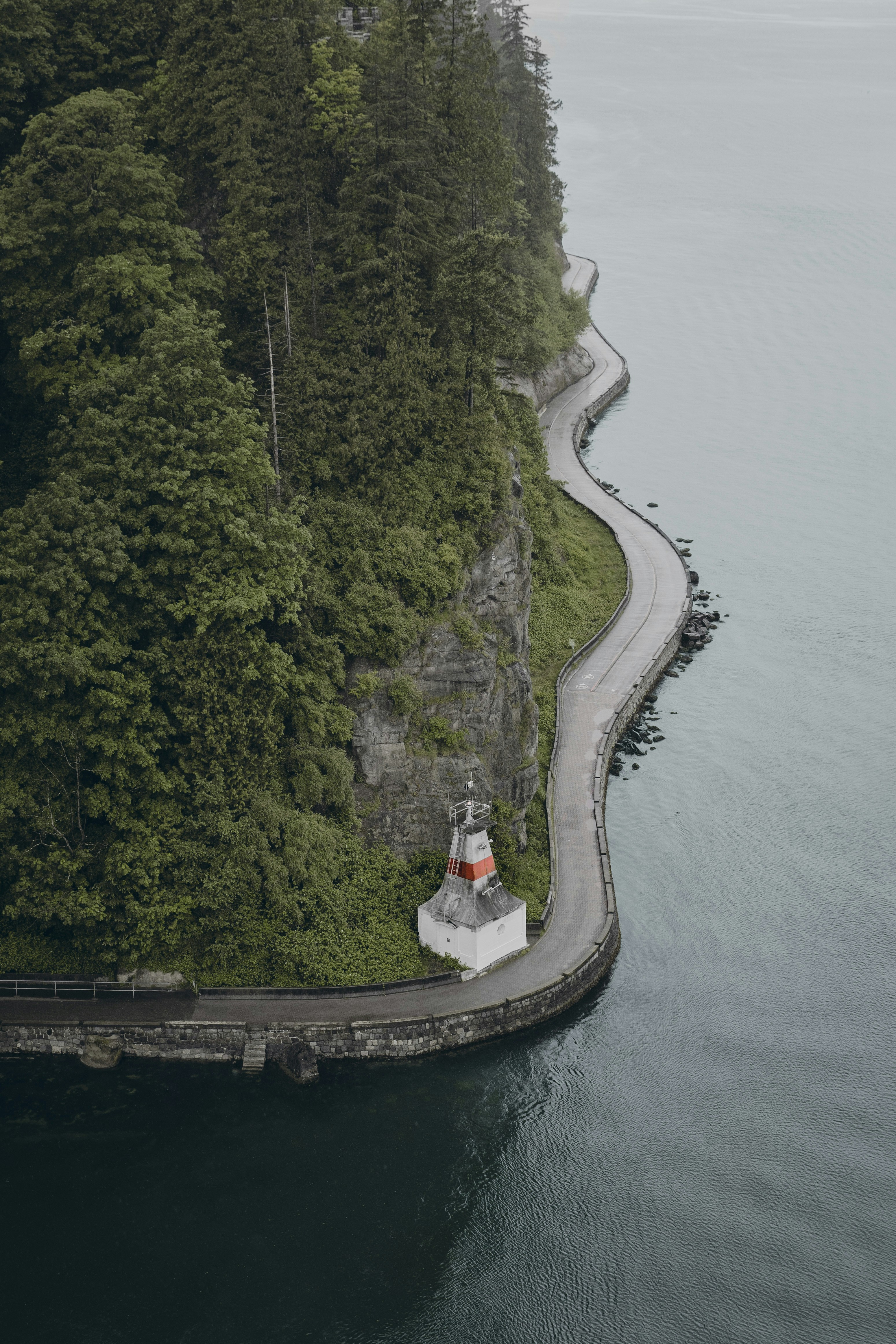 A coastal road curves around a rocky outcrop, featuring a lighthouse at the base surrounded by lush greenery. The tranquil waters reflect the serene landscape.