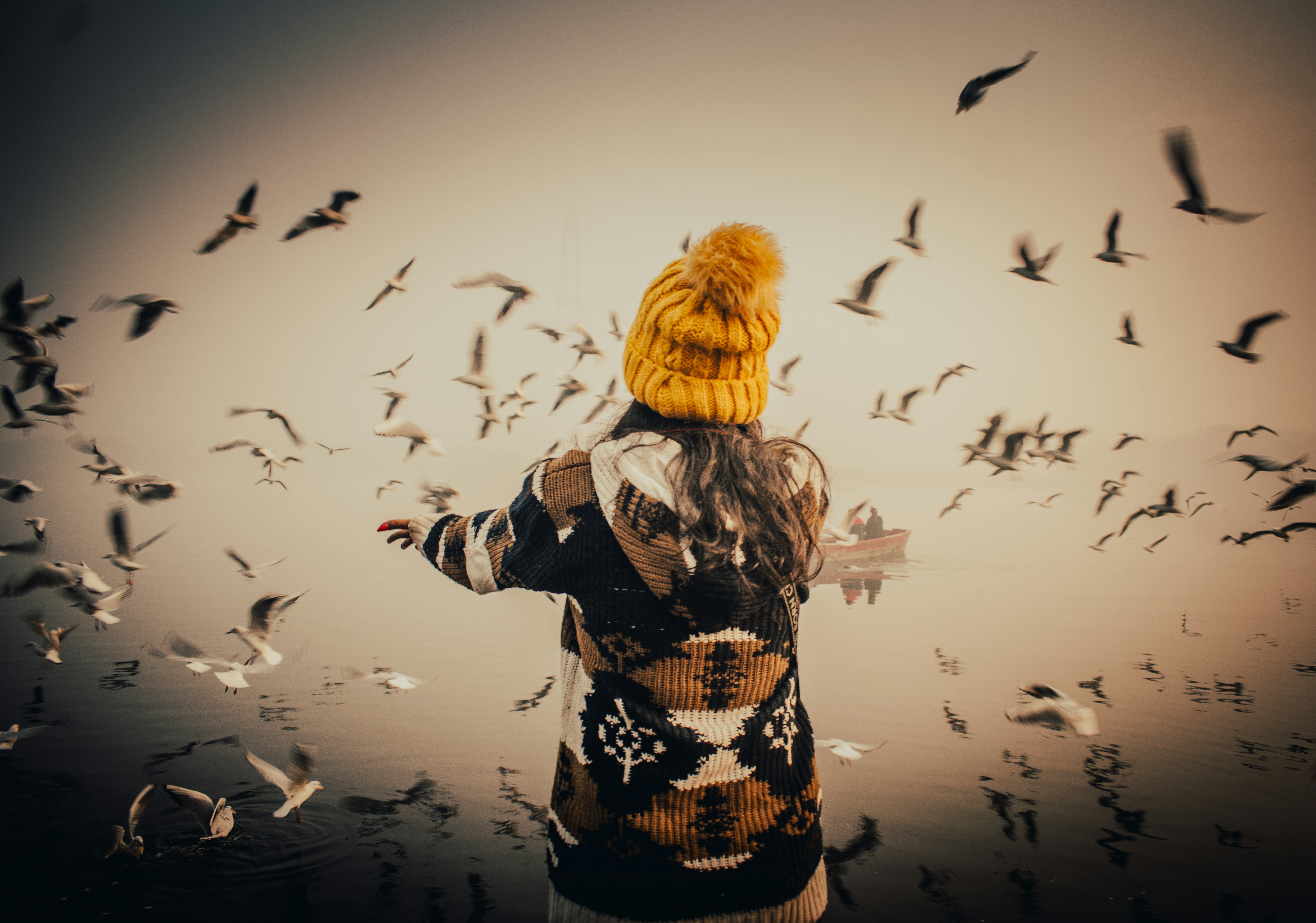 Woman in a patterned coat and yellow hat surrounded by seagulls near a serene lake.
