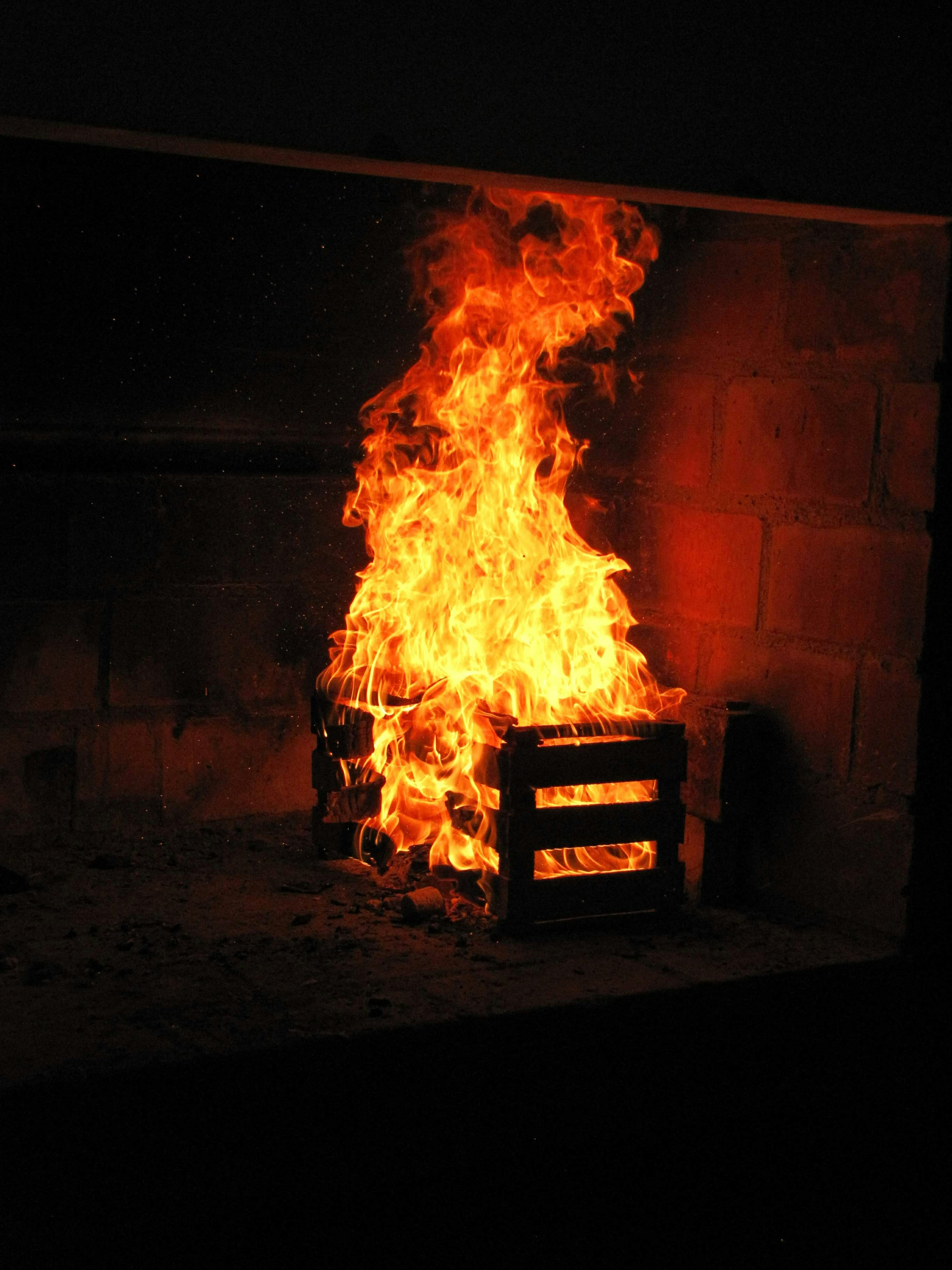 Bright flames engulf wooden crates in a dark fireplace, illuminating the surrounding bricks with a warm glow.
