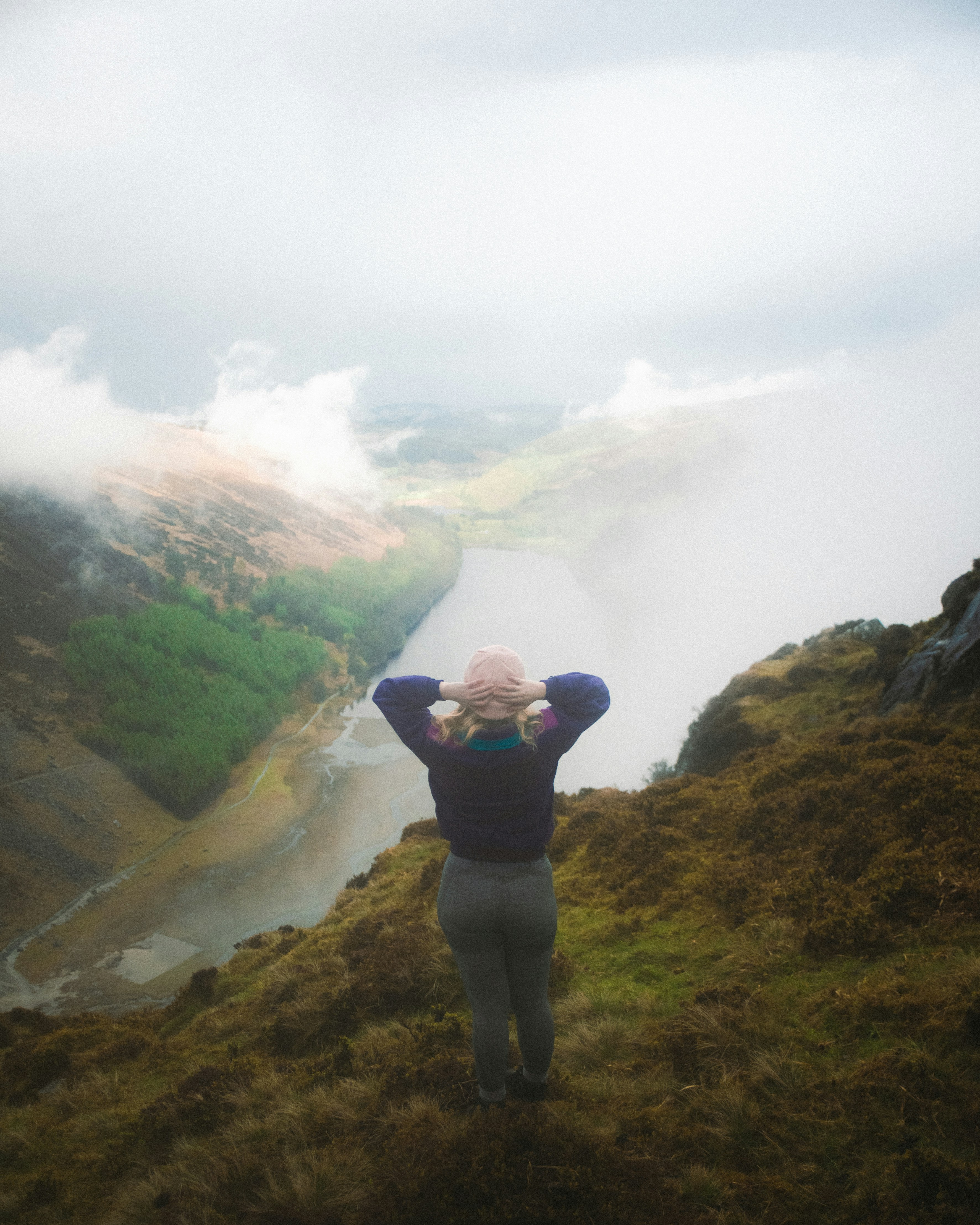 Person standing on a hillside, gazing at a winding river surrounded by lush greenery and misty mountains. The scene captures a moment of reflection in a serene landscape.