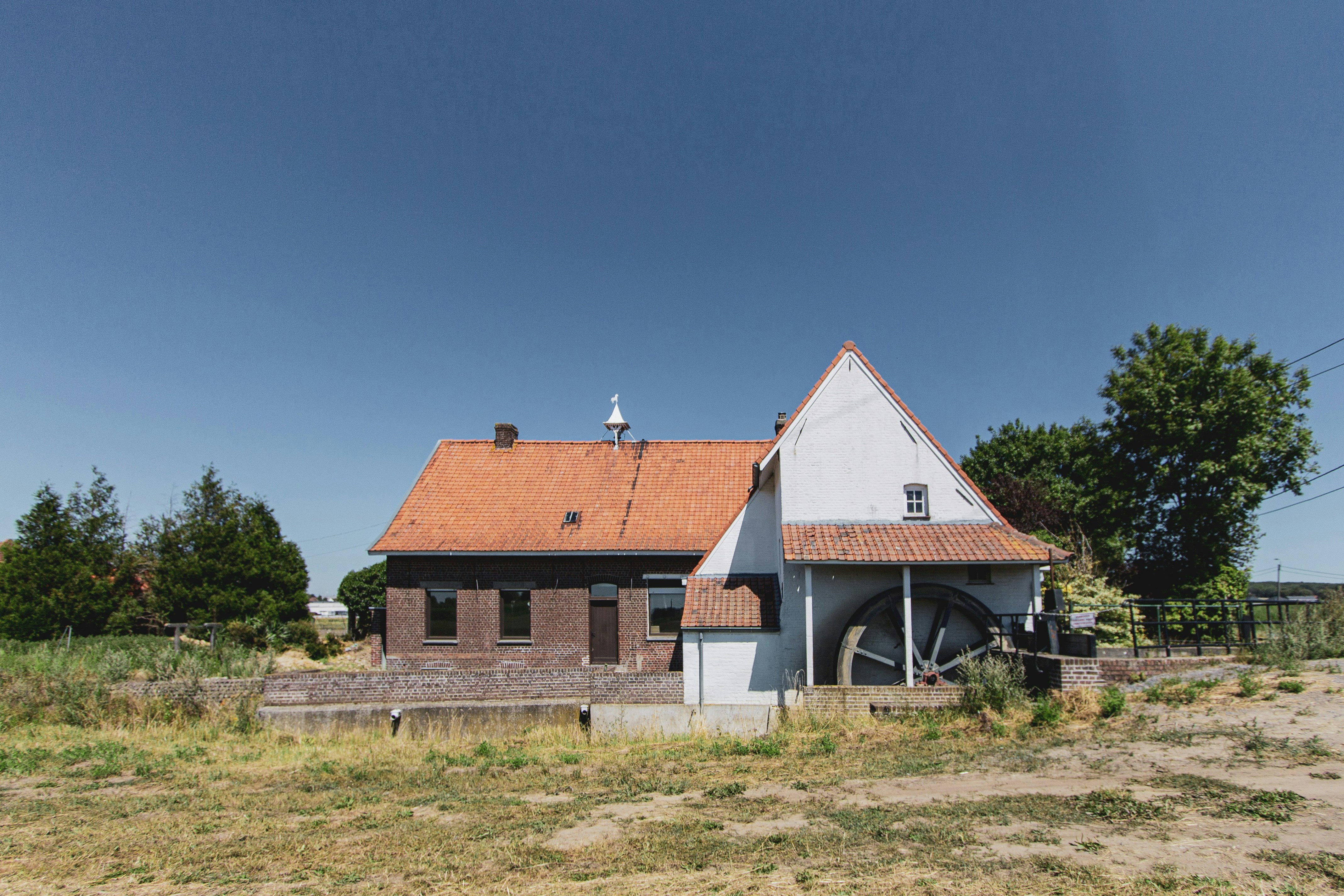 A quaint, historic building with a distinctive wheel, surrounded by lush greenery and a clear blue sky. The structure reflects traditional craftsmanship and rural charm.