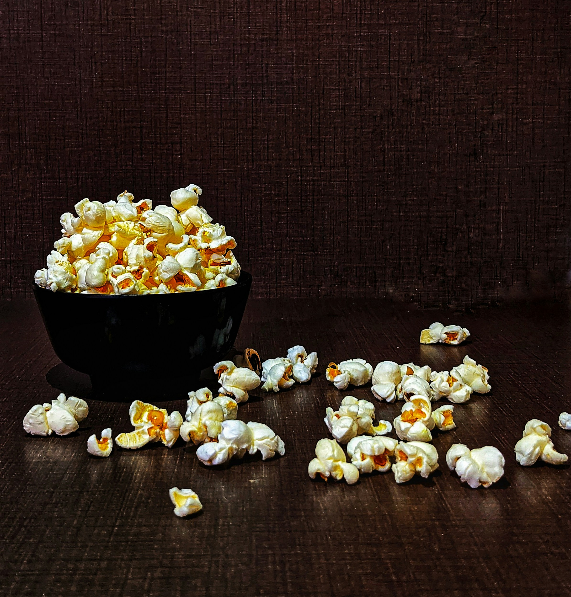 A black bowl overflowing with freshly popped popcorn, surrounded by scattered kernels on a dark wooden surface.