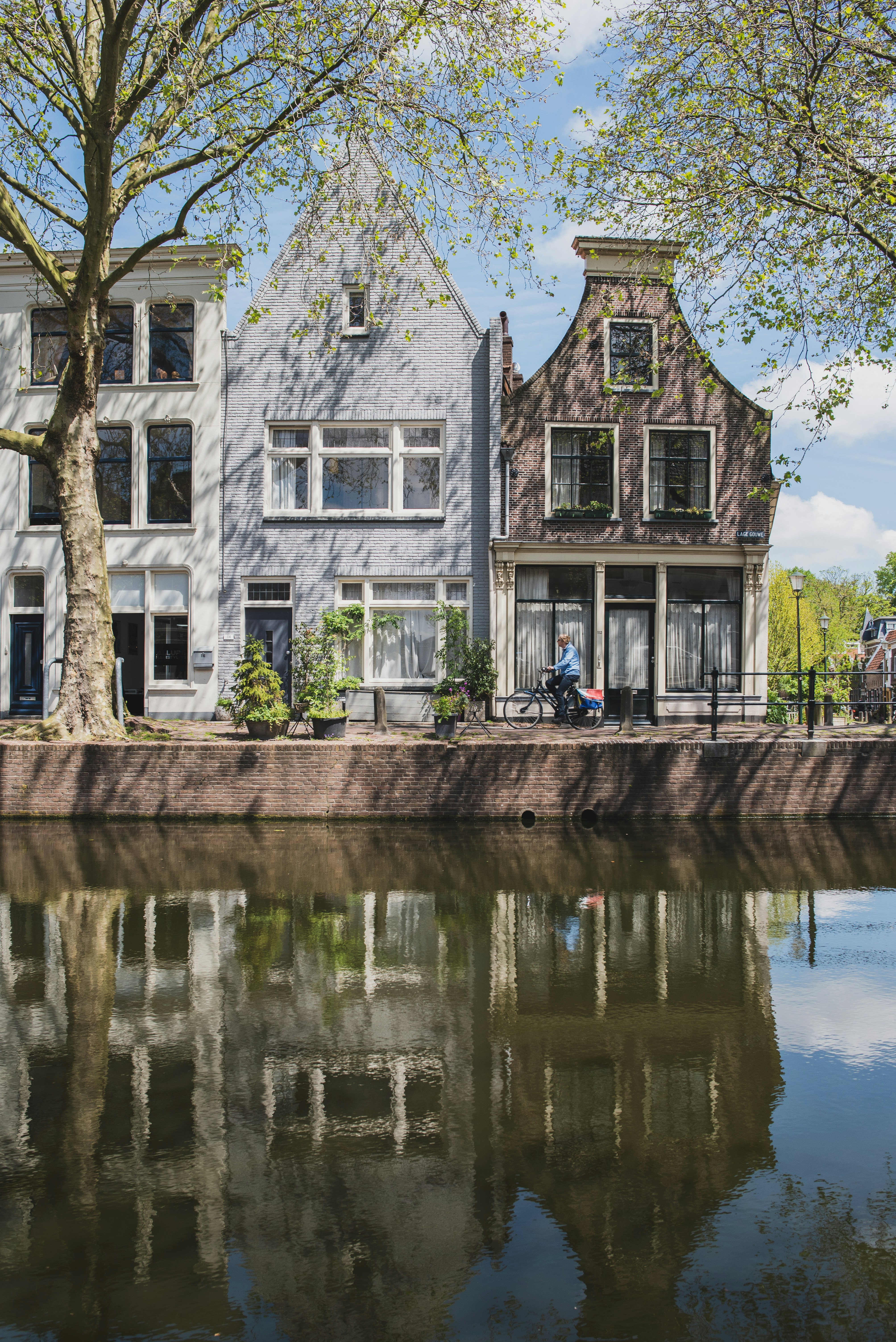 Charming canal-side houses reflecting in the water, framed by lush trees and a clear sky. A cyclist pauses, adding life to the serene scene.