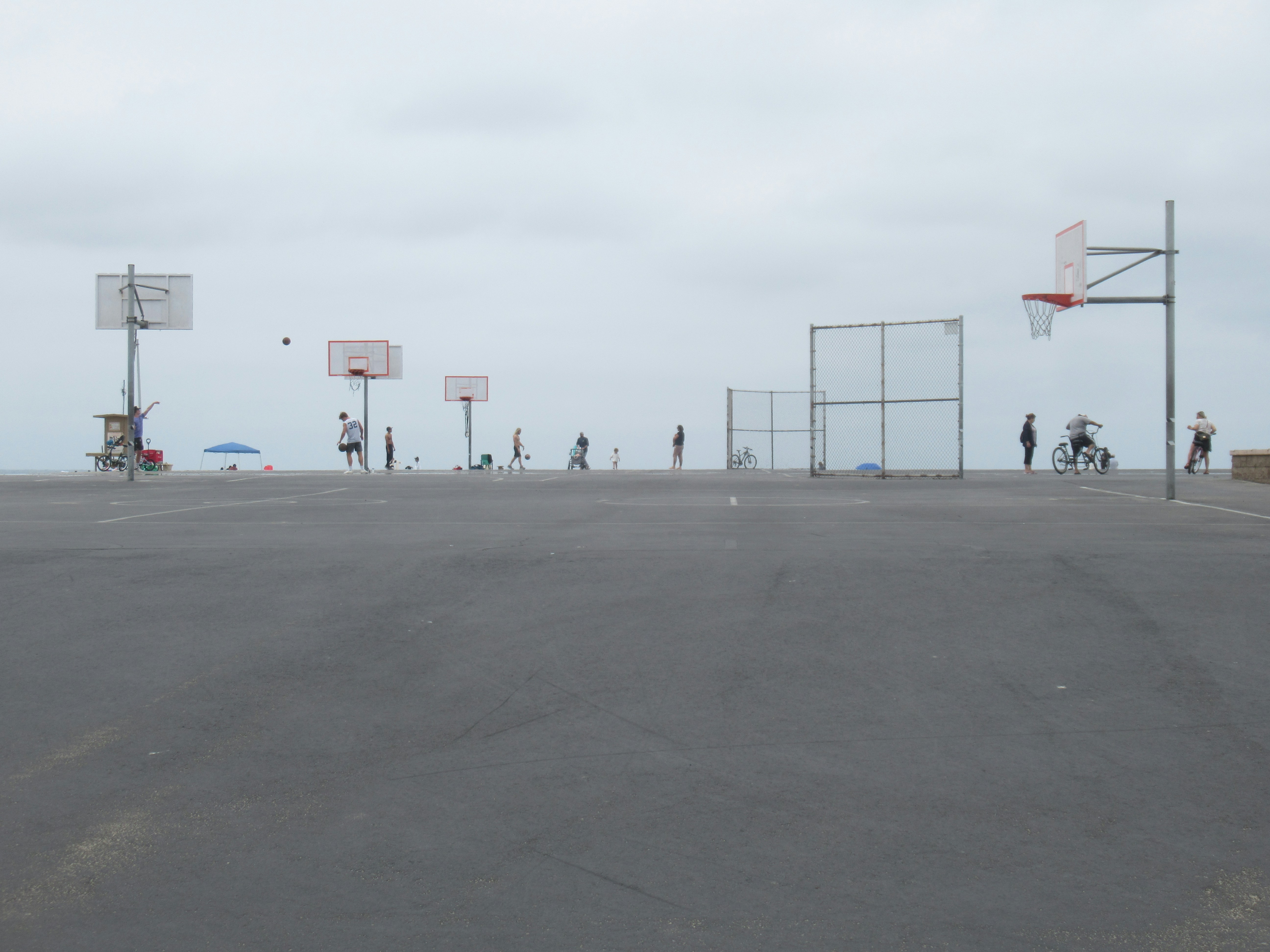 Distant basketball players punctuate a wide, empty boardwalk court under an overcast sky. This photograph emphasizes the row of hoops and the open horizon.