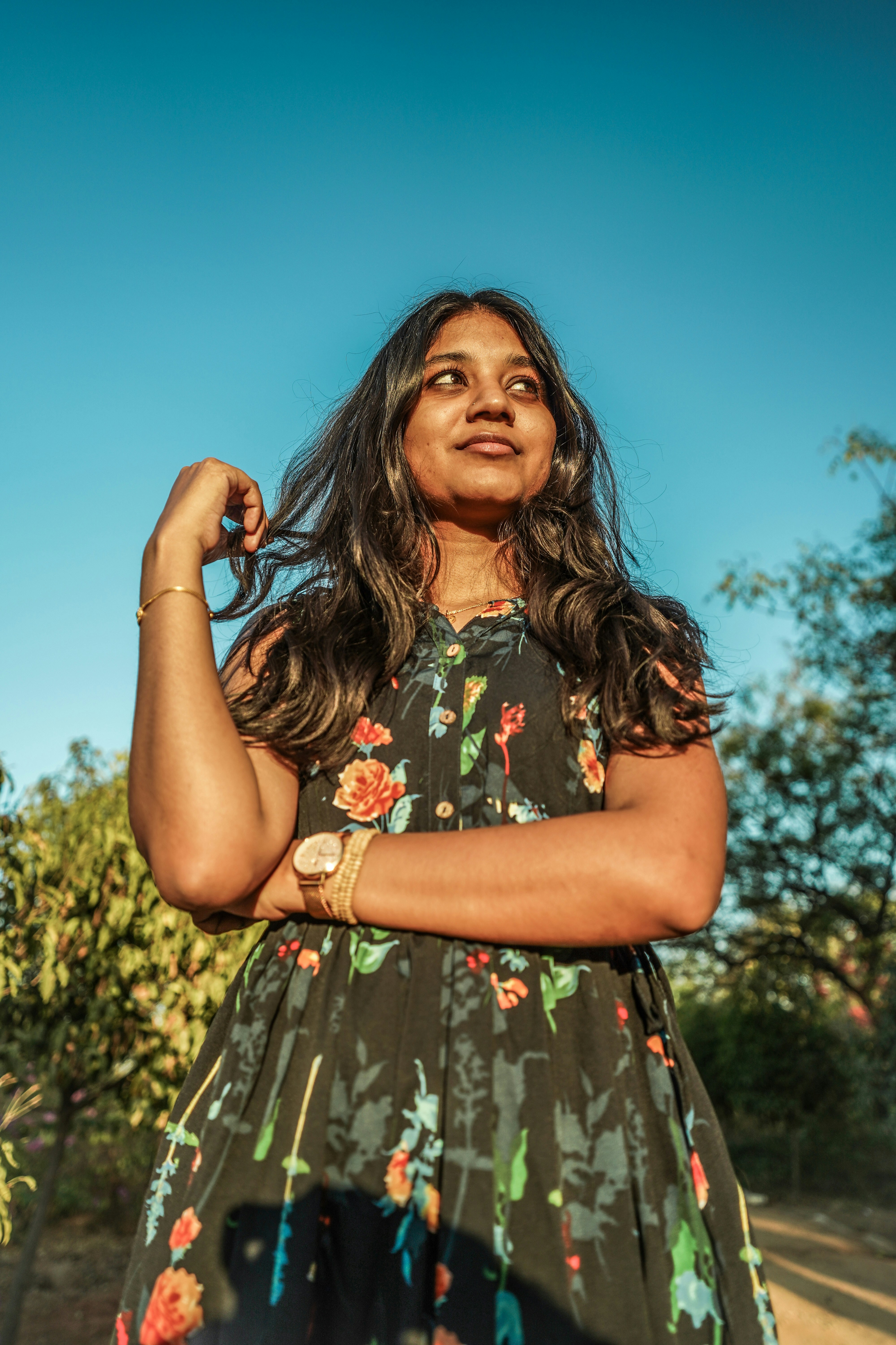 A woman in a floral dress stands confidently against a clear blue sky, her hair illuminated by the warm glow of the setting sun.