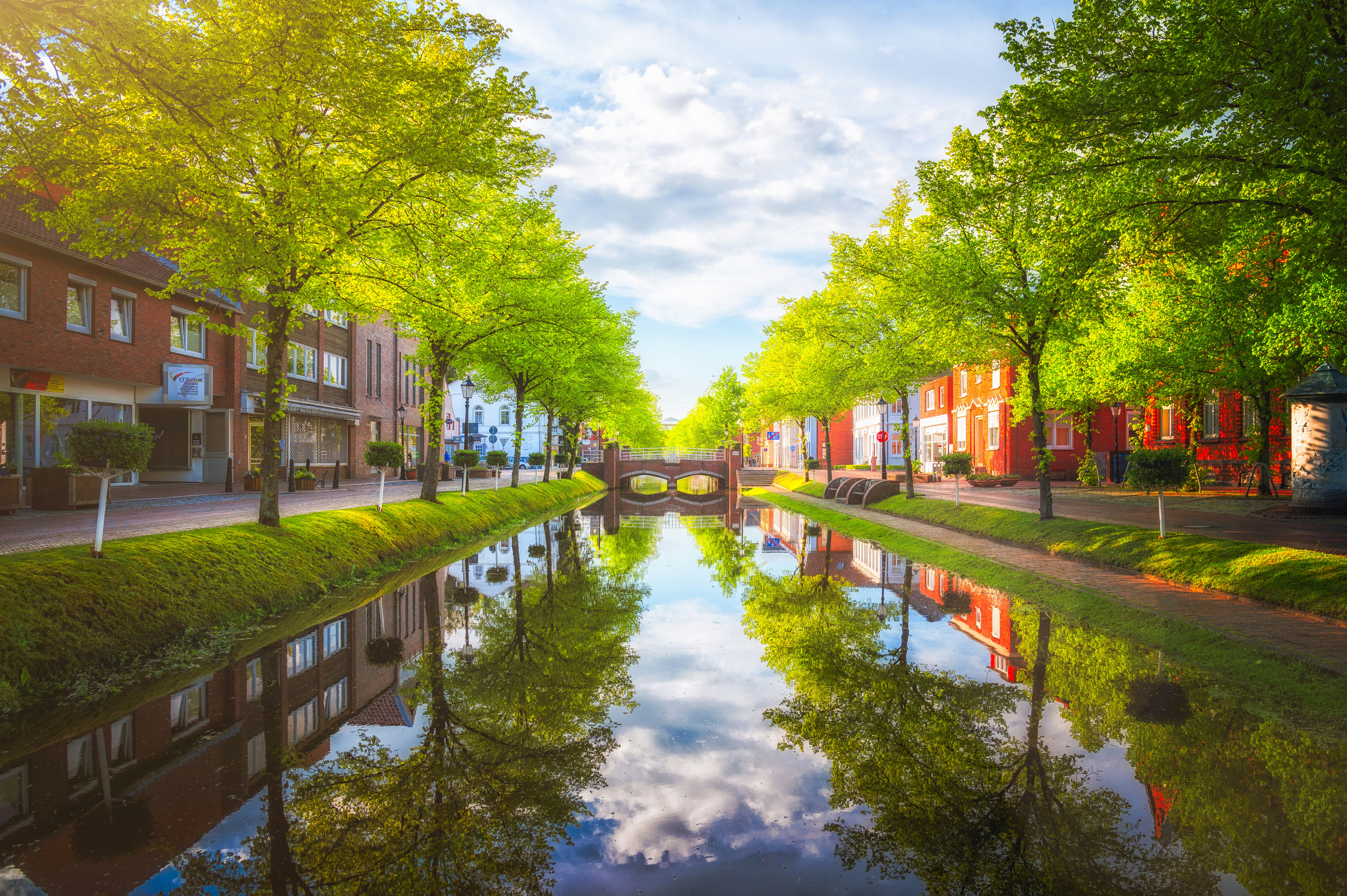 Tree-lined canal reflecting vibrant houses and a stone bridge under a partly cloudy sky.