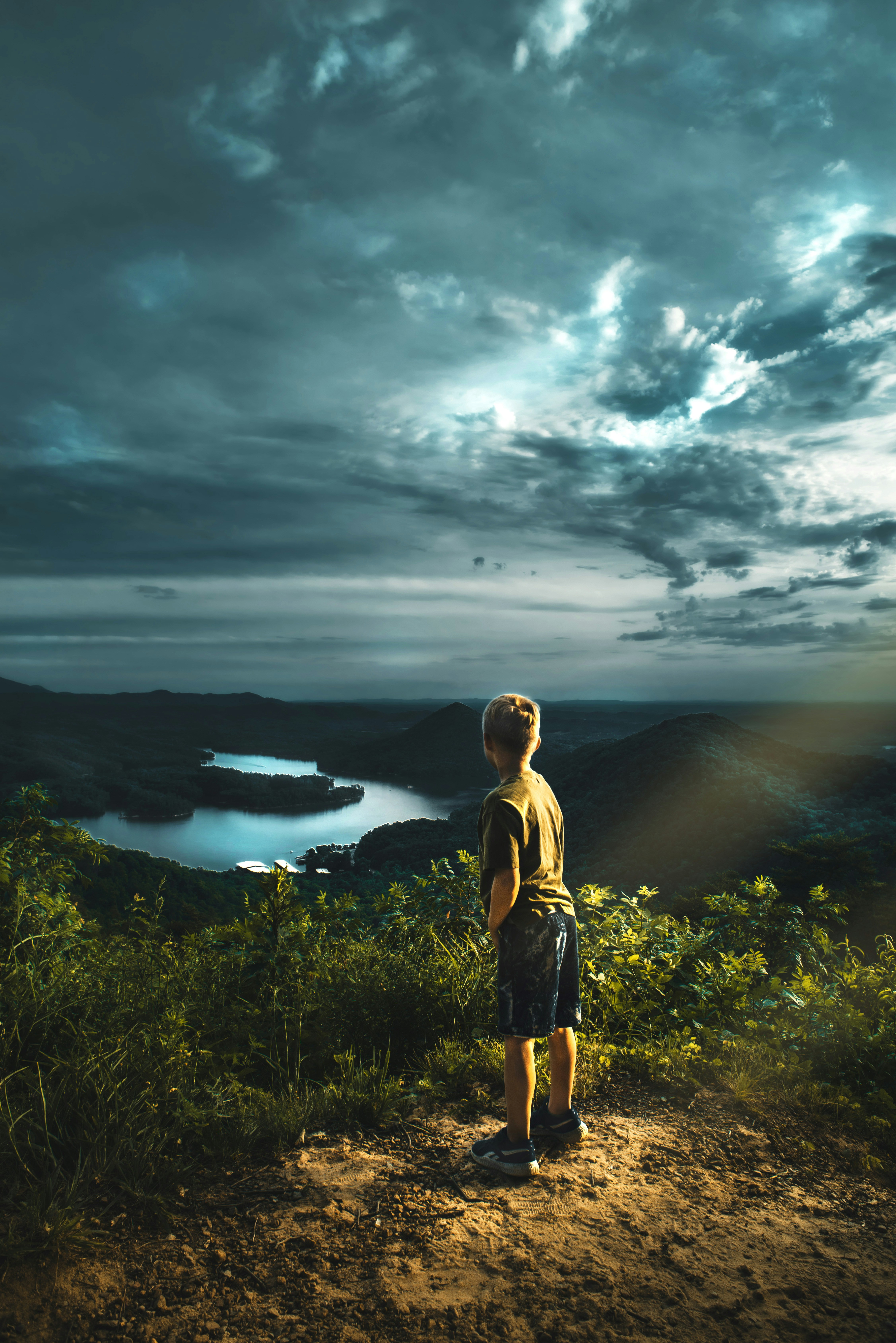 Child standing on a hill overlooking a serene lake under a dramatic evening sky.