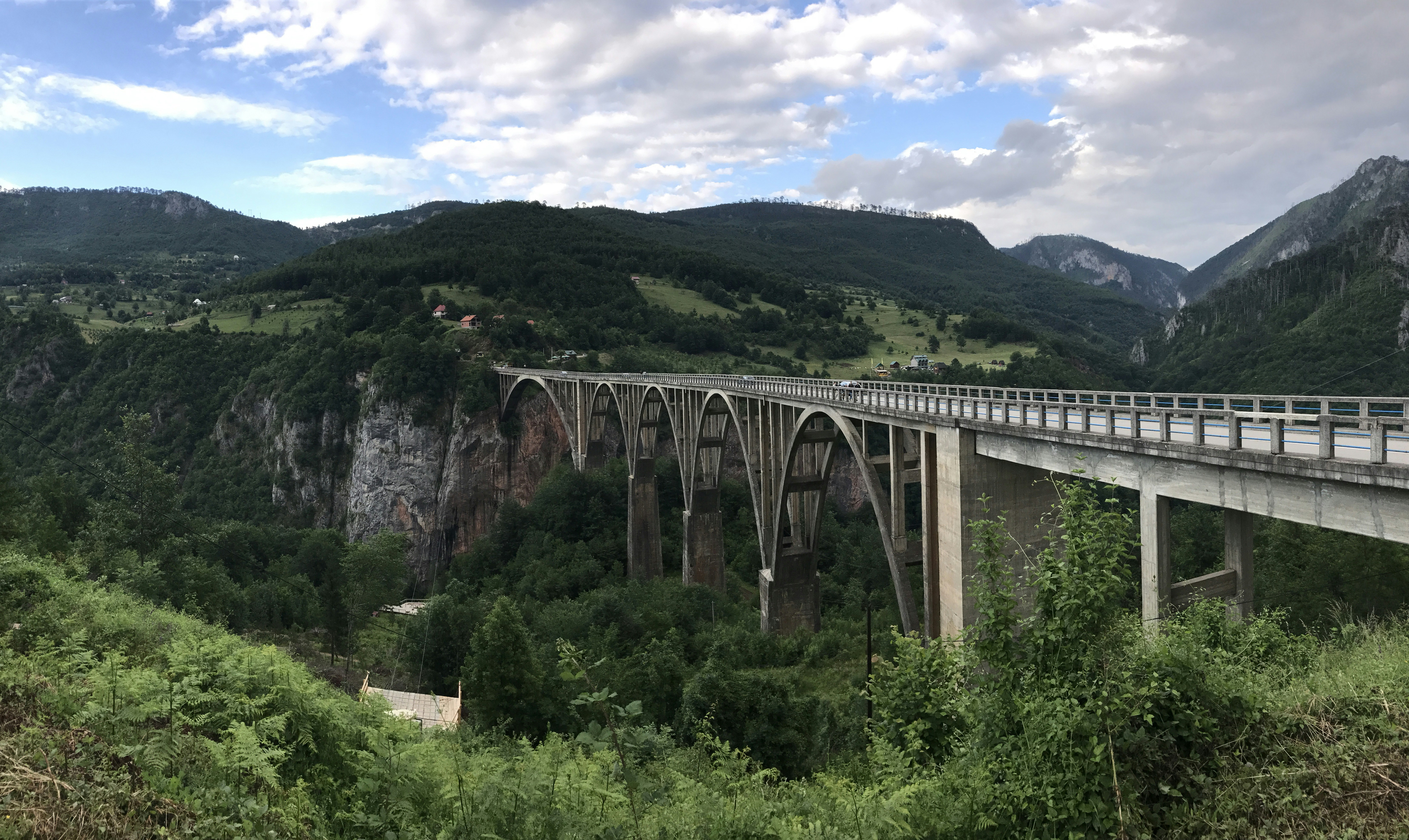 A modern bridge arches gracefully over a lush valley, framed by rolling hills and dramatic cliffs. The scene captures a blend of human engineering and natural beauty.