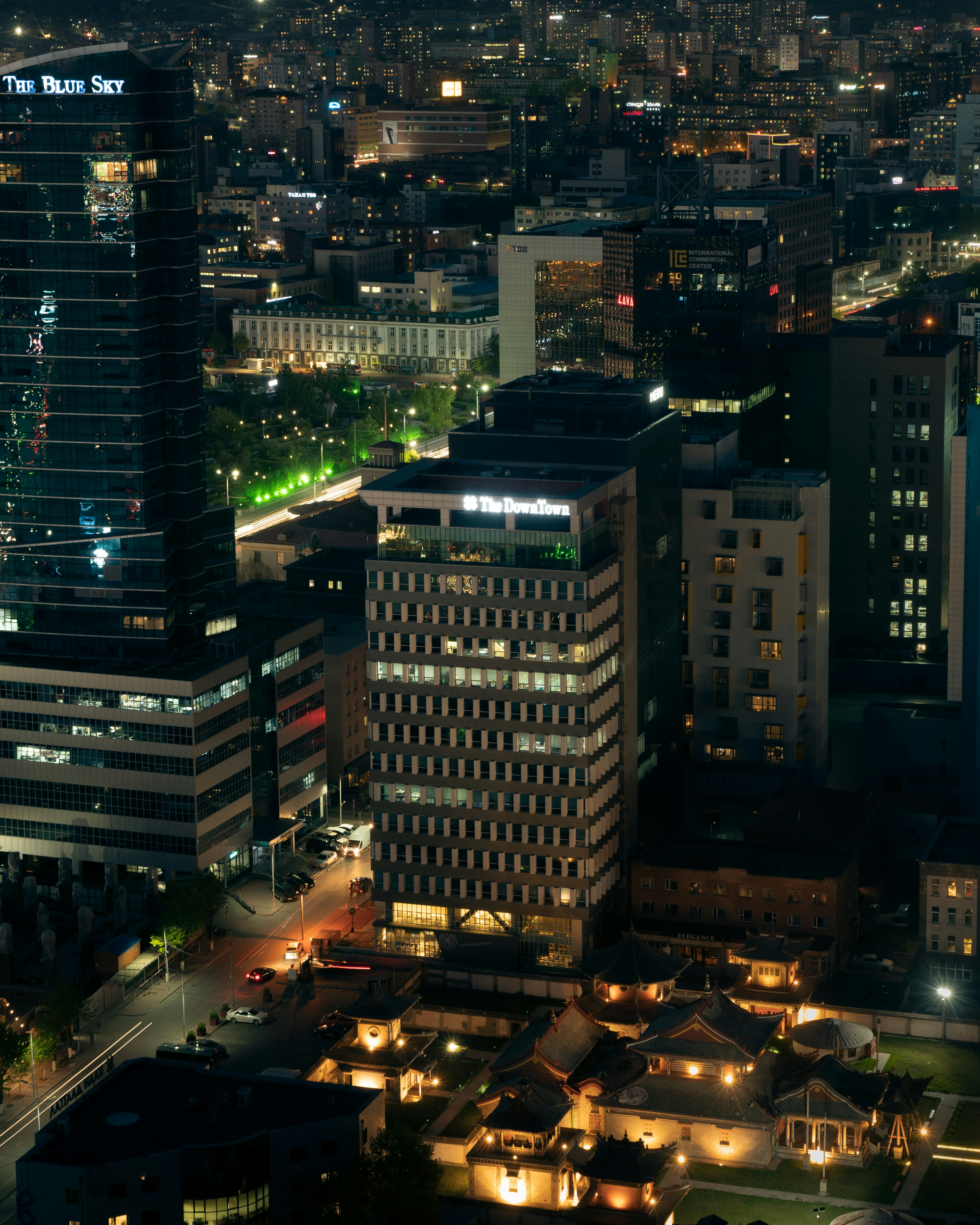 A vibrant cityscape at night showcasing illuminated buildings and bustling streets, with traditional architecture nestled below modern structures.