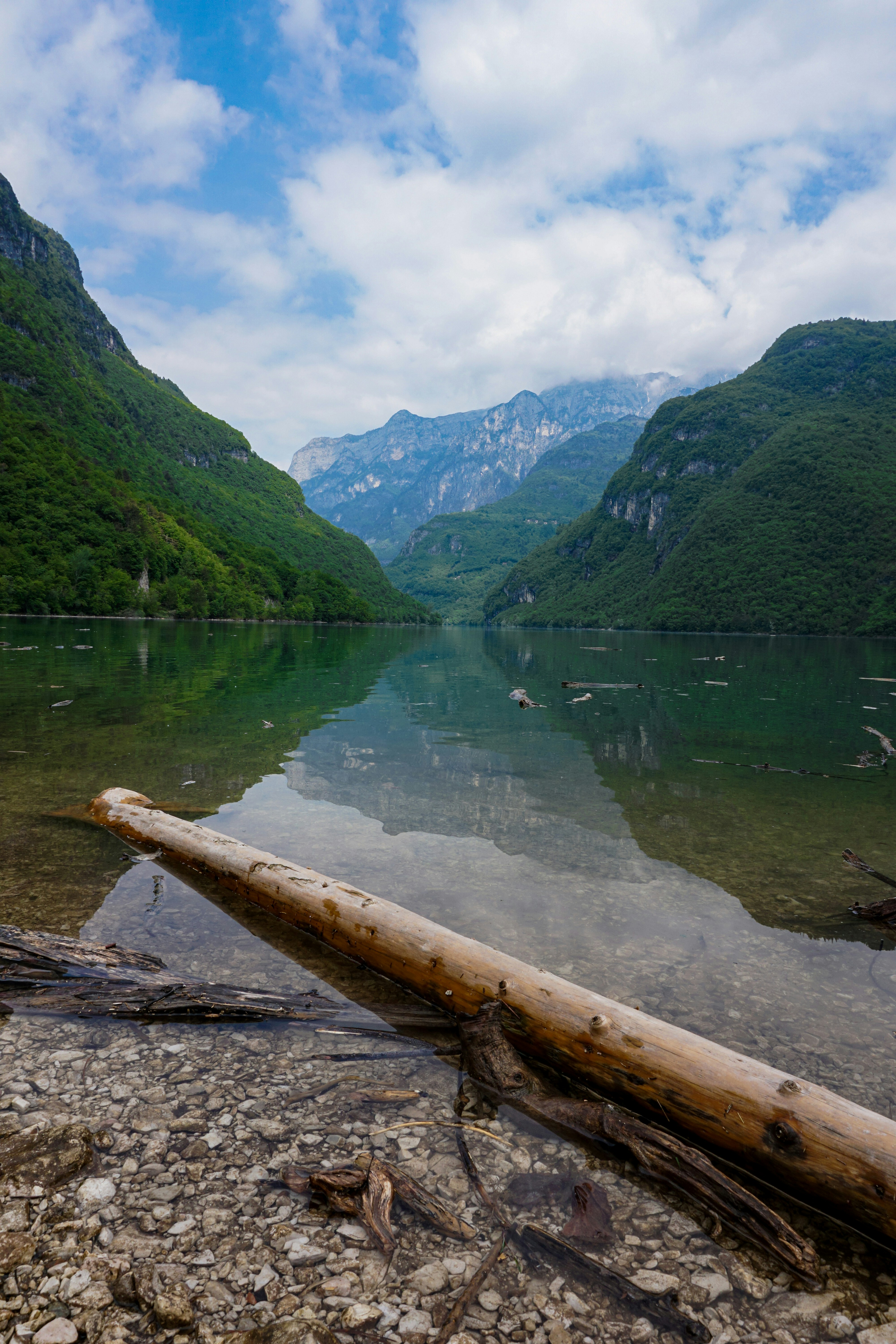A tranquil lake reflecting the surrounding mountains, with a fallen log and pebbly shoreline in the foreground.
