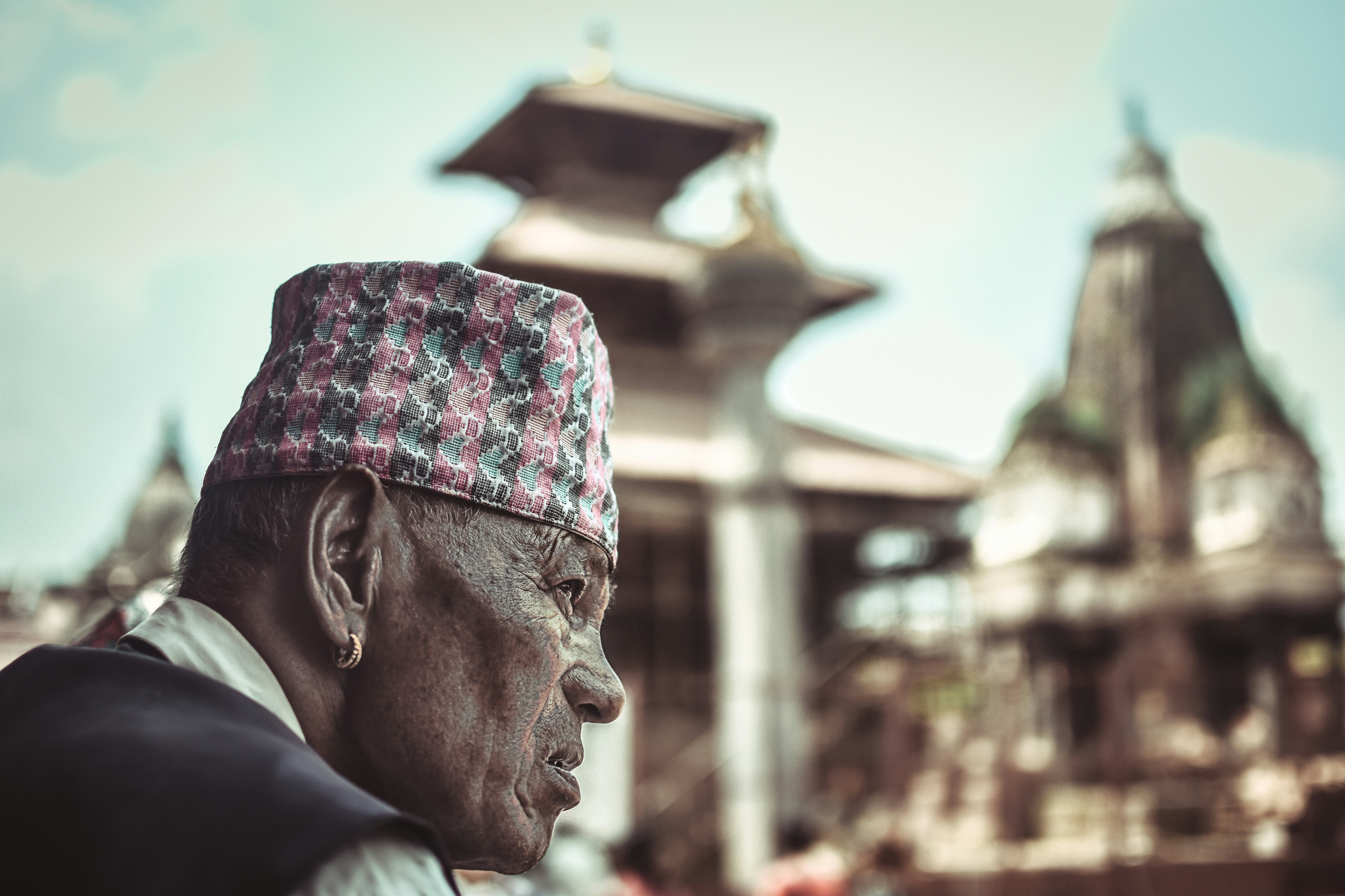 Elderly man wearing a traditional patterned hat with blurred ancient temples in the background.