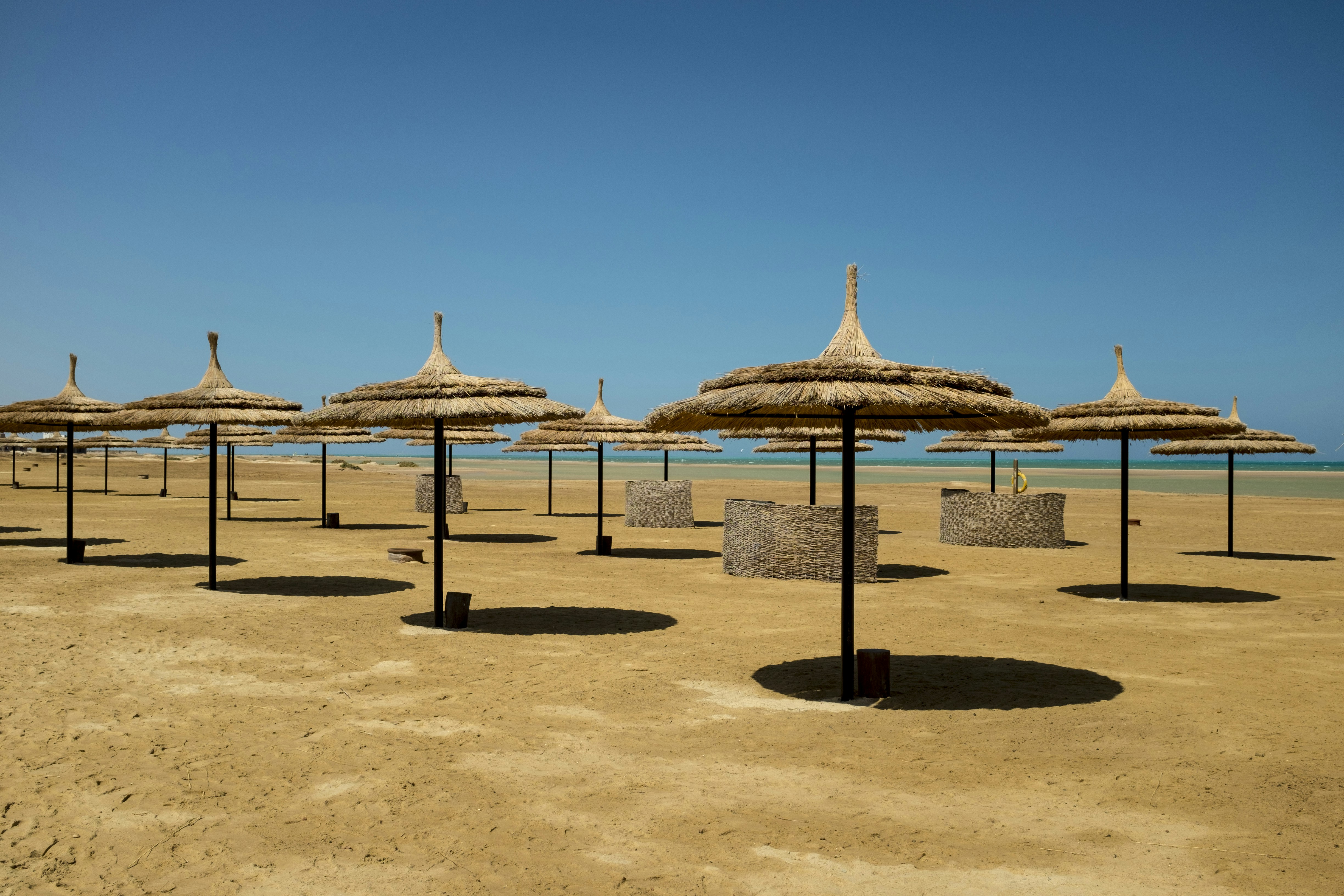 A row of straw umbrellas casting shadows on a sunlit sandy beach, with calm turquoise waters in the background.