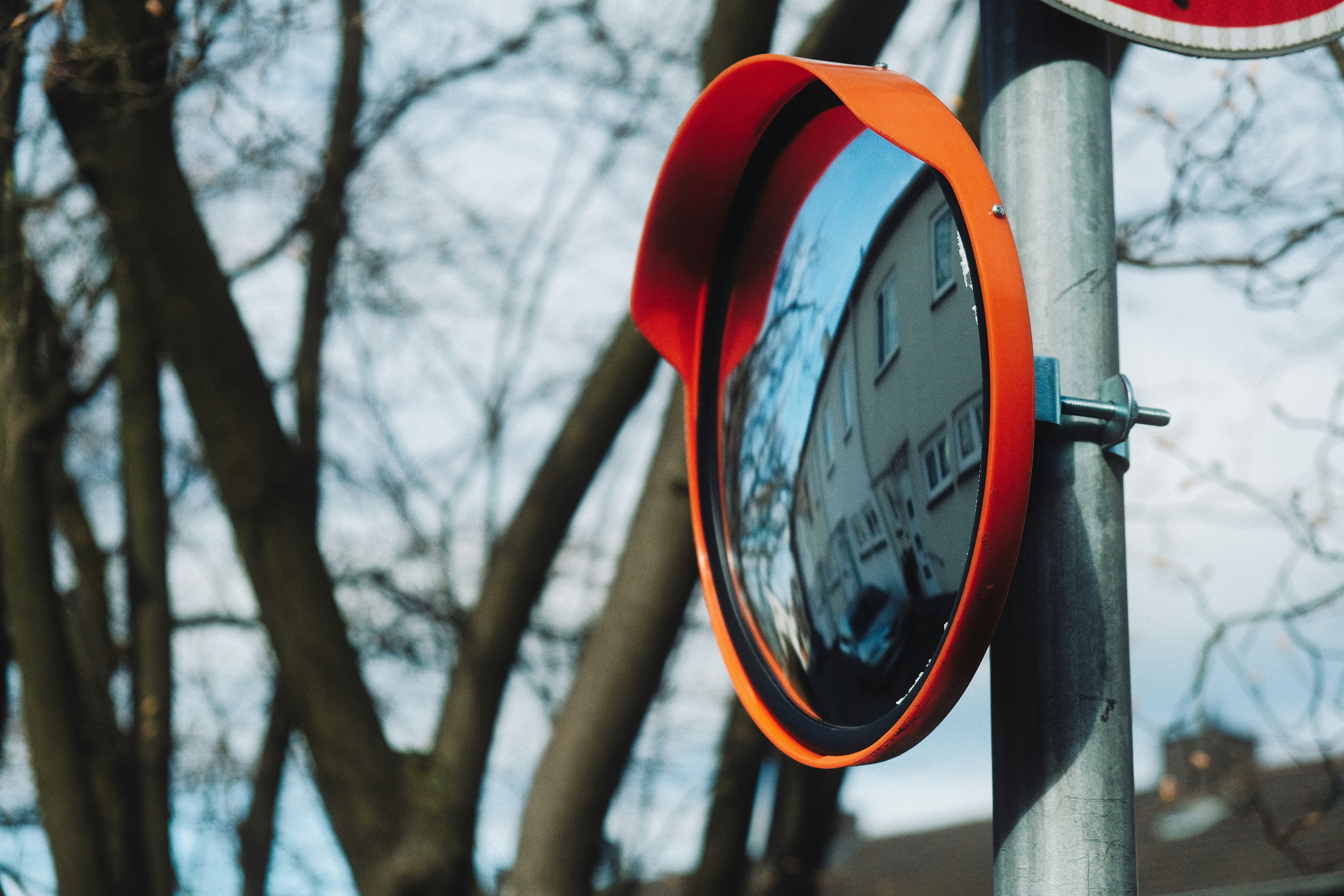 Convex traffic mirror reflecting bare trees and a building under a cloudy sky.