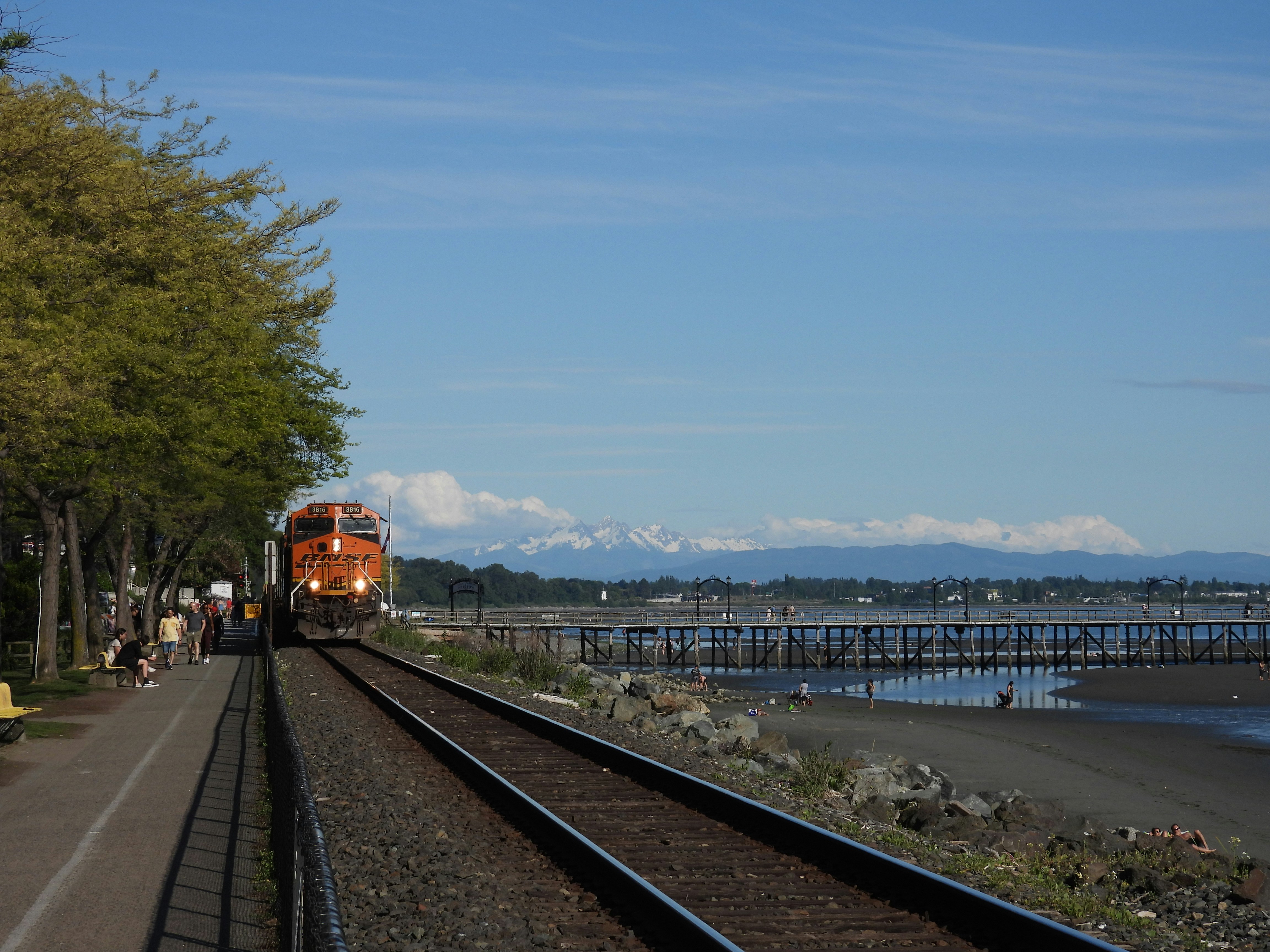 Orange locomotive sits on coastal tracks along a pedestrian promenade, with people gathered to the left. A wooden pier extends into calm water on the right, creating a seaside day scene.