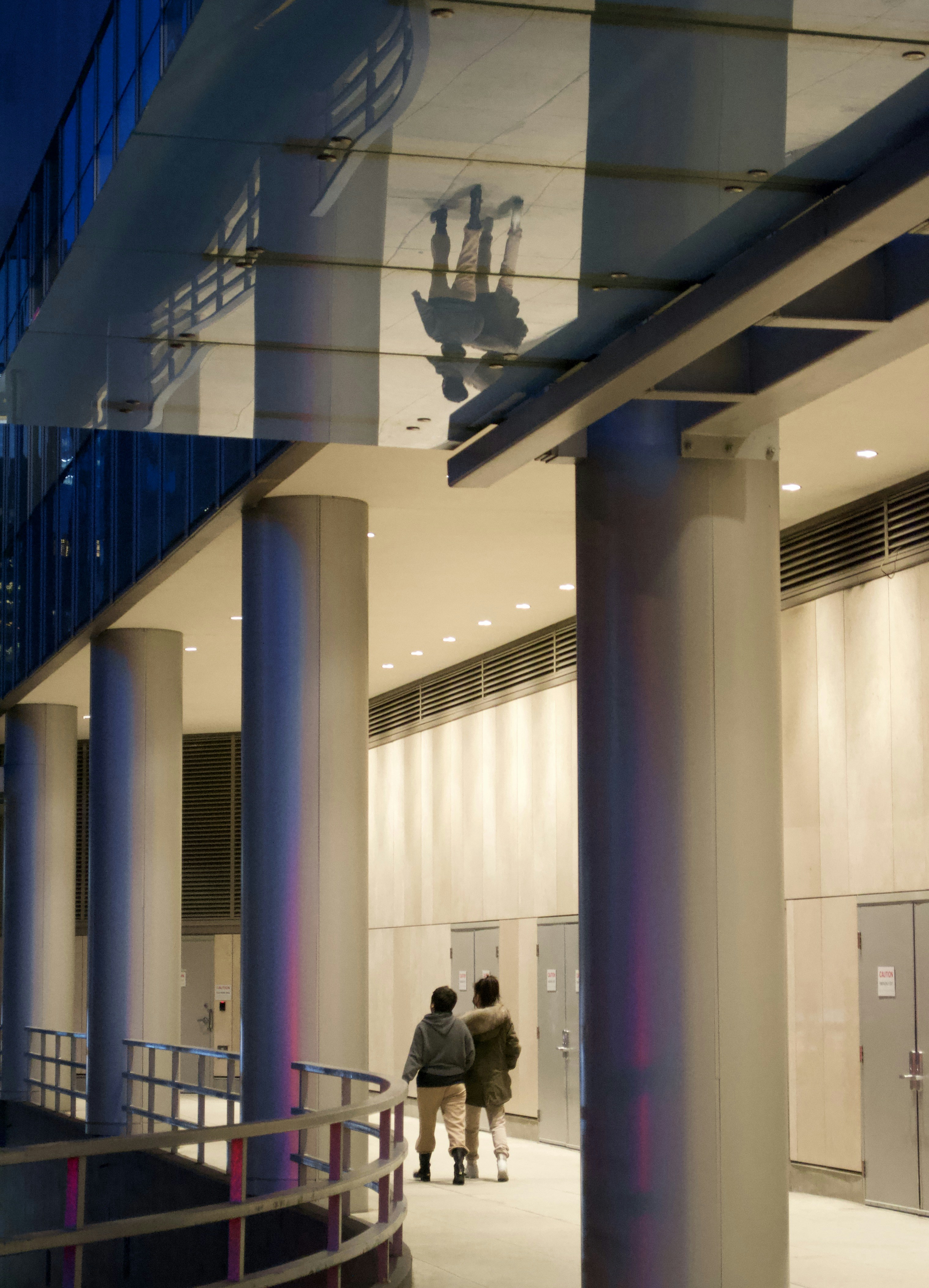 Couple walking beneath a modern architectural overhang, their reflection visible on the glass surface above. Soft lighting enhances the urban ambiance.