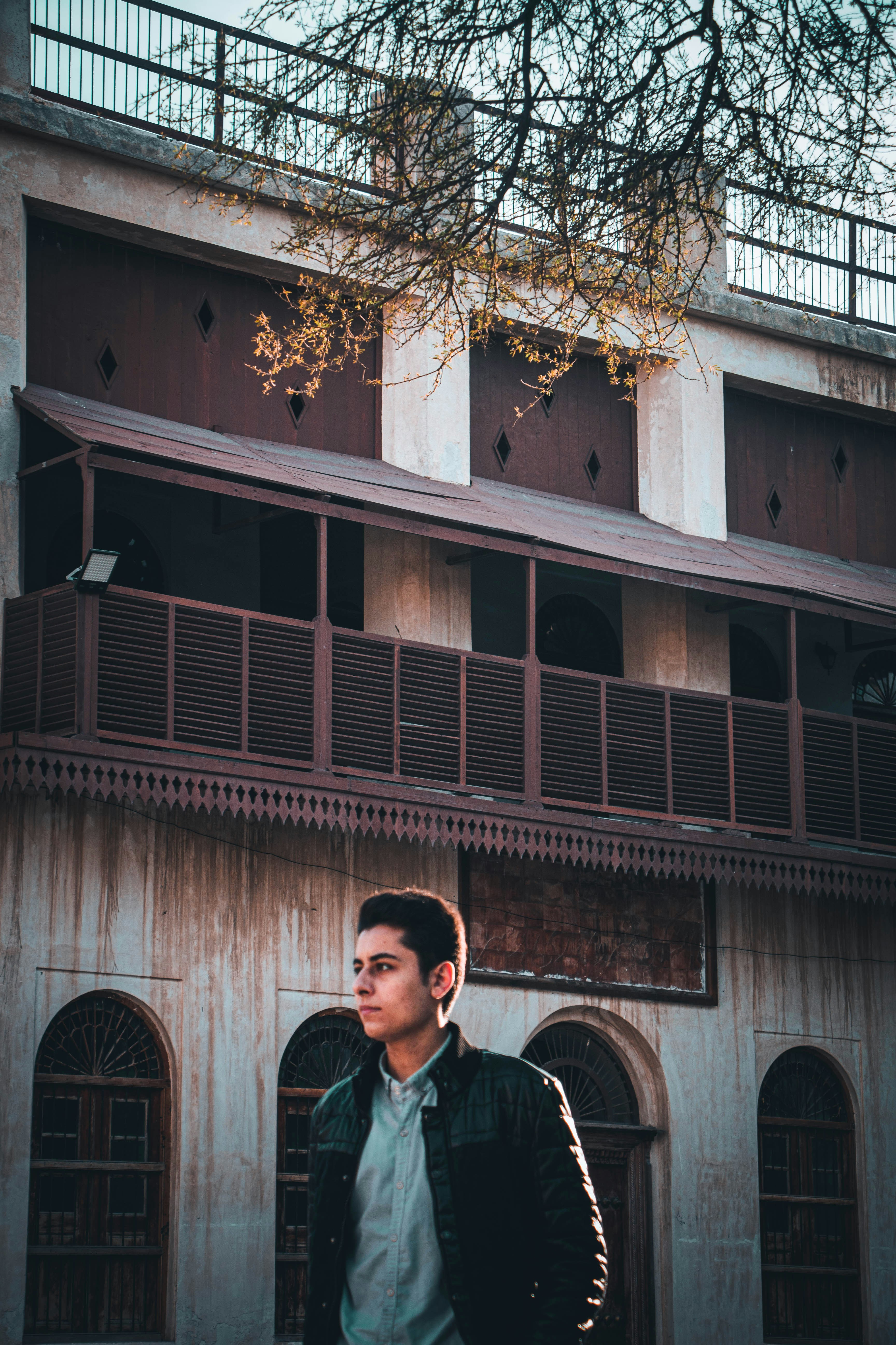 Young man in a leather jacket stands in front of a traditional building with ornate windows and a balcony. The scene blends contemporary style with historical architecture.