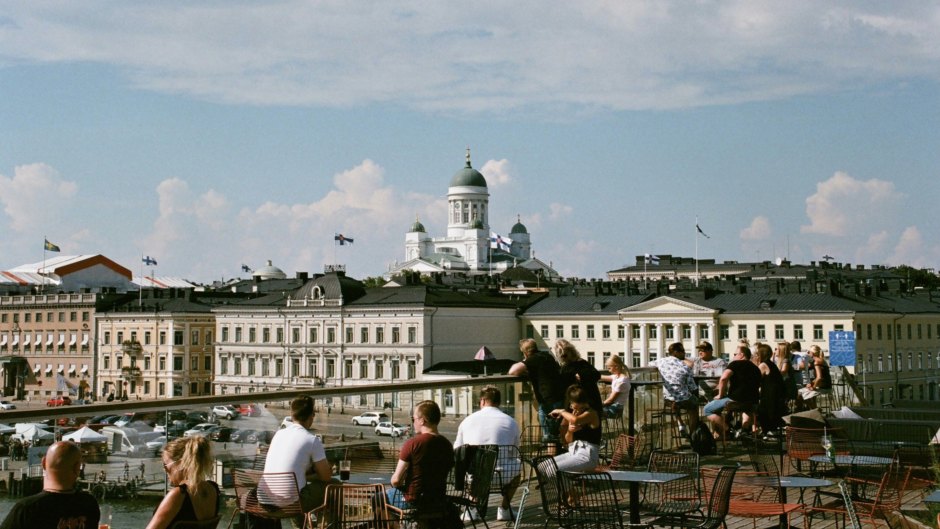 Busy waterfront plaza with outdoor seating and pedestrians, backed by a row of historic buildings. A domed, green-roofed city hall rises at center beneath a clear blue sky.