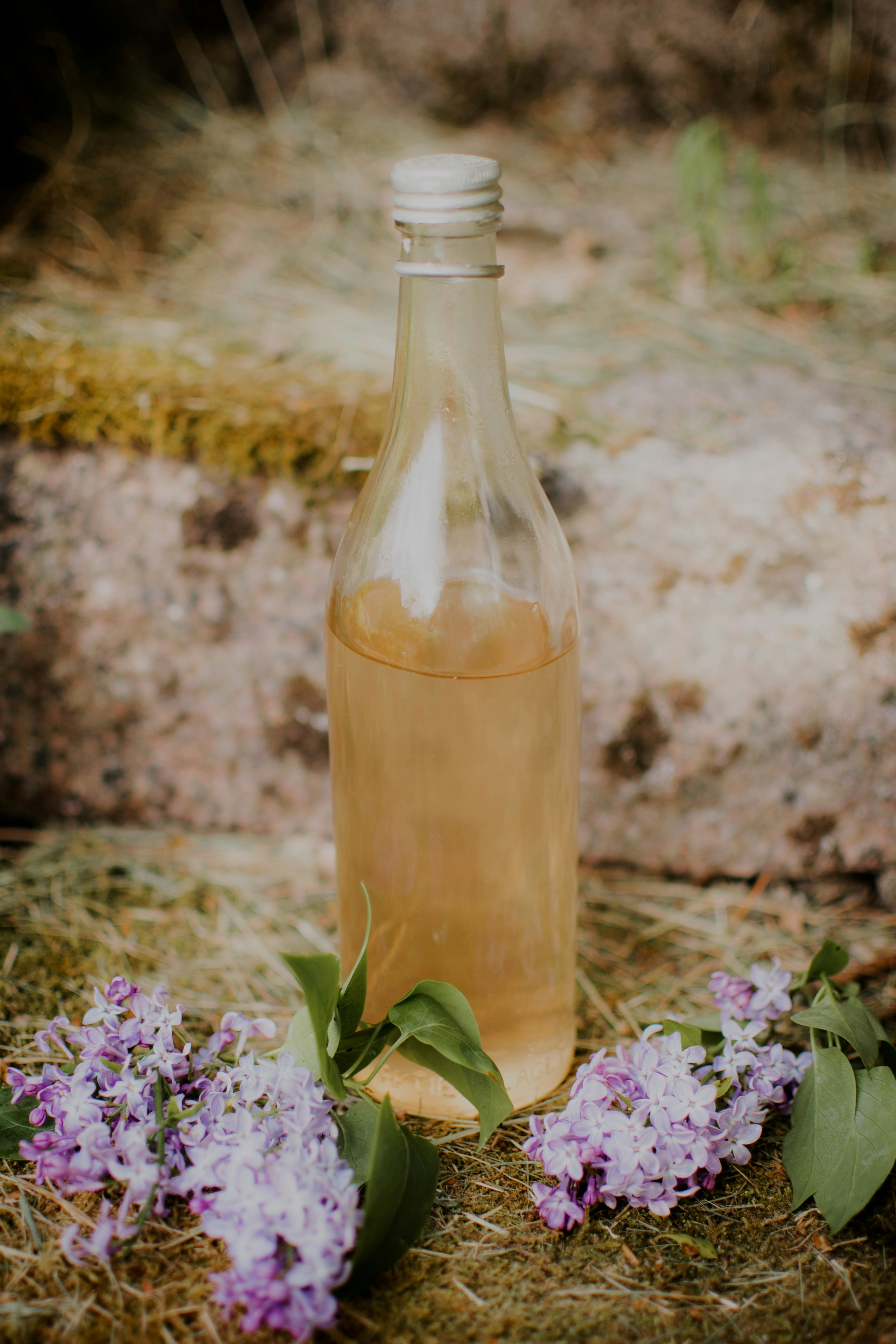 A glass bottle filled with light-colored liquid rests on a mossy surface, surrounded by blooming lilacs and green leaves.
