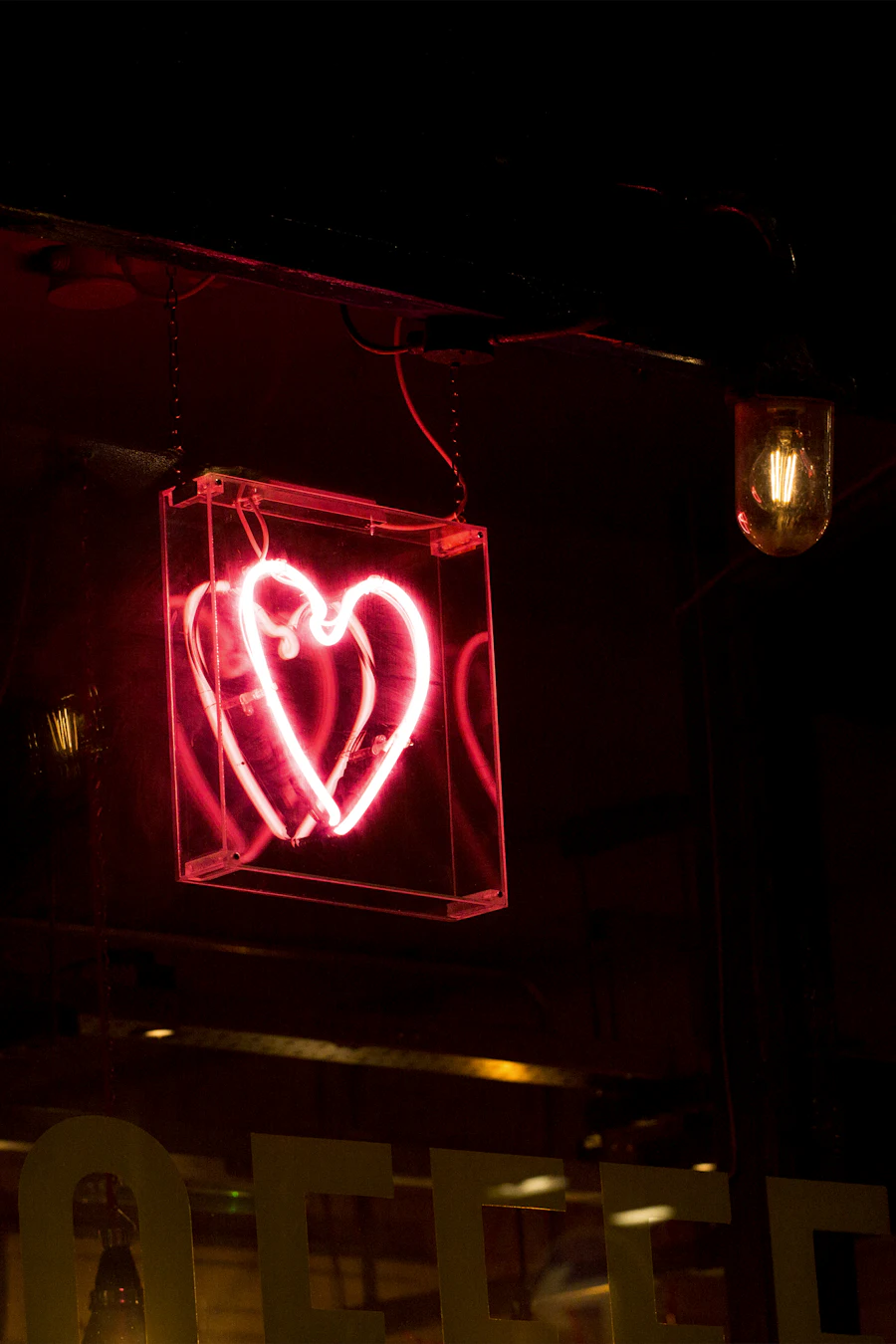 Bright red neon heart glowing inside a clear rectangular frame against a dark interior, with a few warm bulbs nearby and partial lettering along the bottom. The scene feels moody and urban.