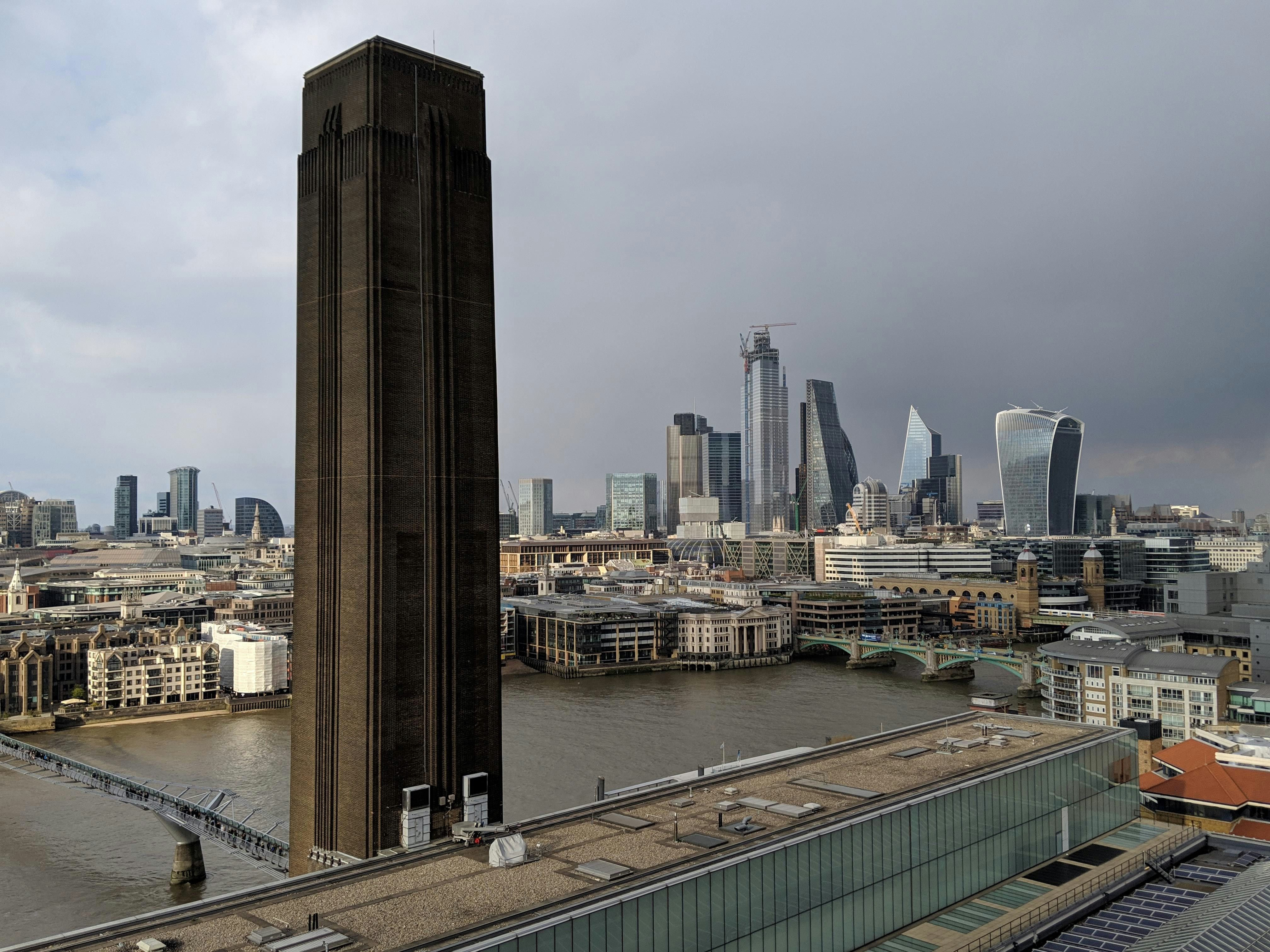 The imposing silhouette of the Tate Modern tower contrasts with the sleek lines of London's financial district in a dynamic urban landscape.