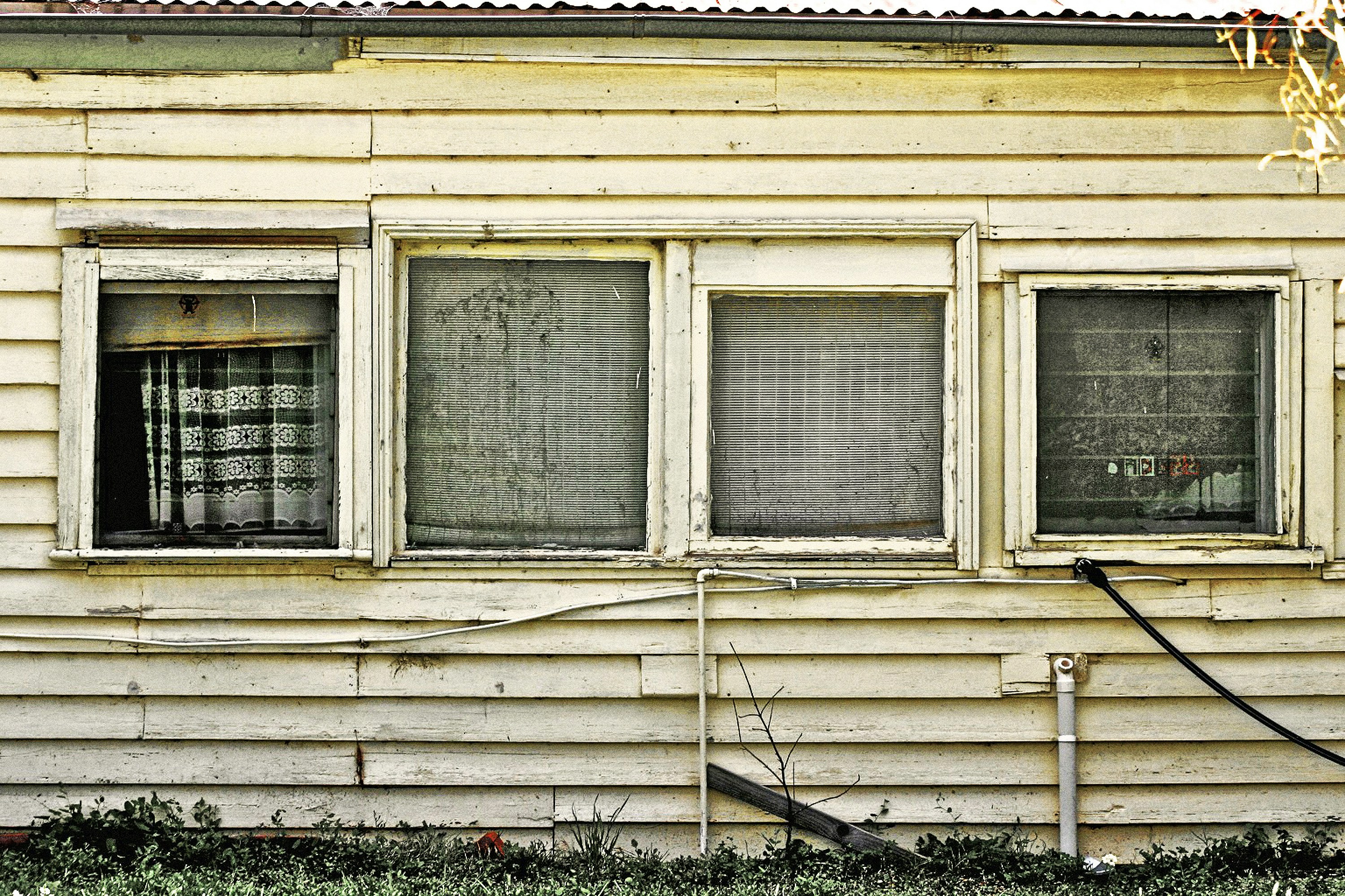 Three weathered windows of an old house, showcasing peeling paint and faded curtains, hinting at stories untold.