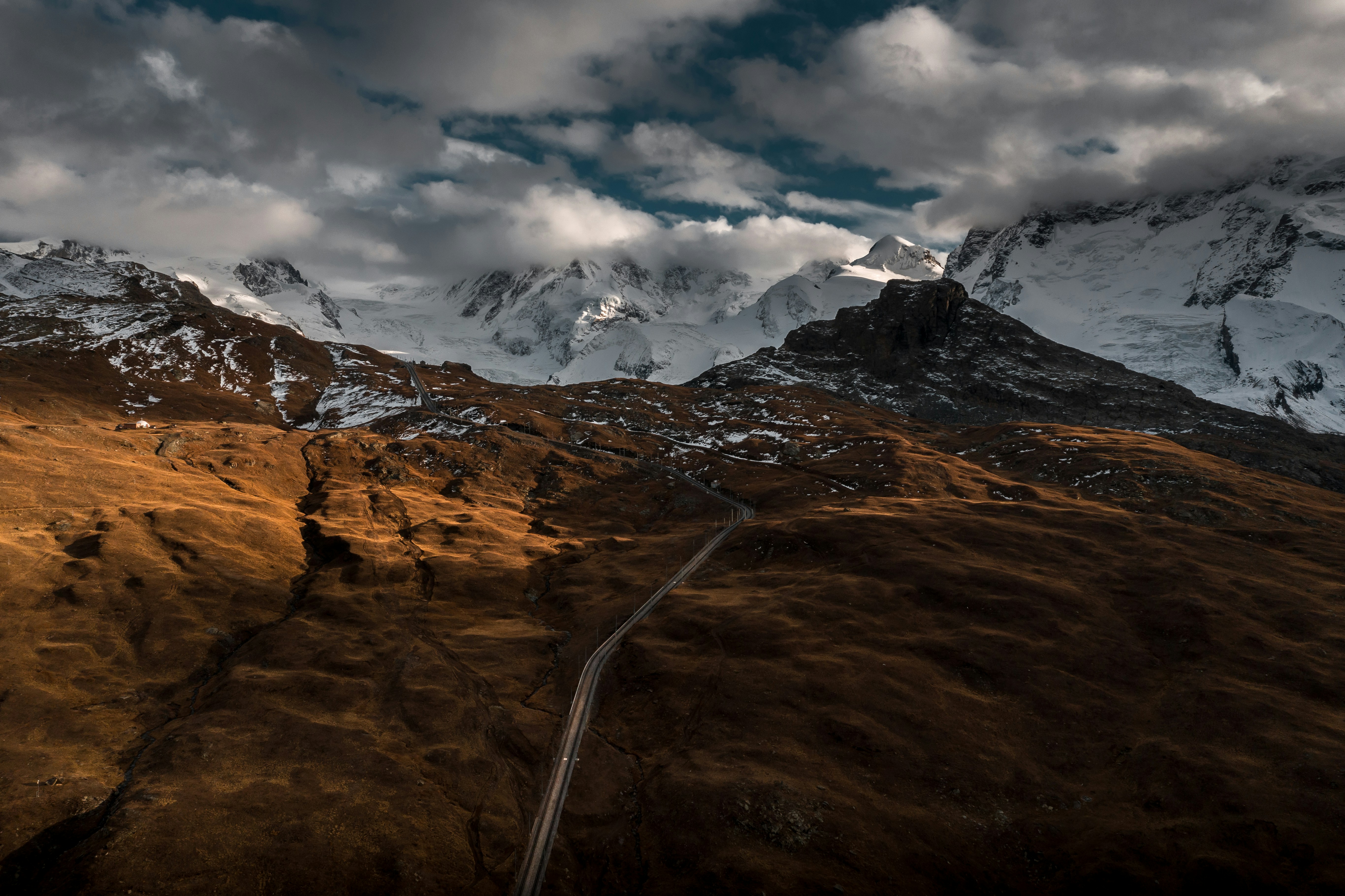 Winding road cuts through rugged alpine landscape, flanked by dramatic snow-capped peaks and contrasting golden terrain.